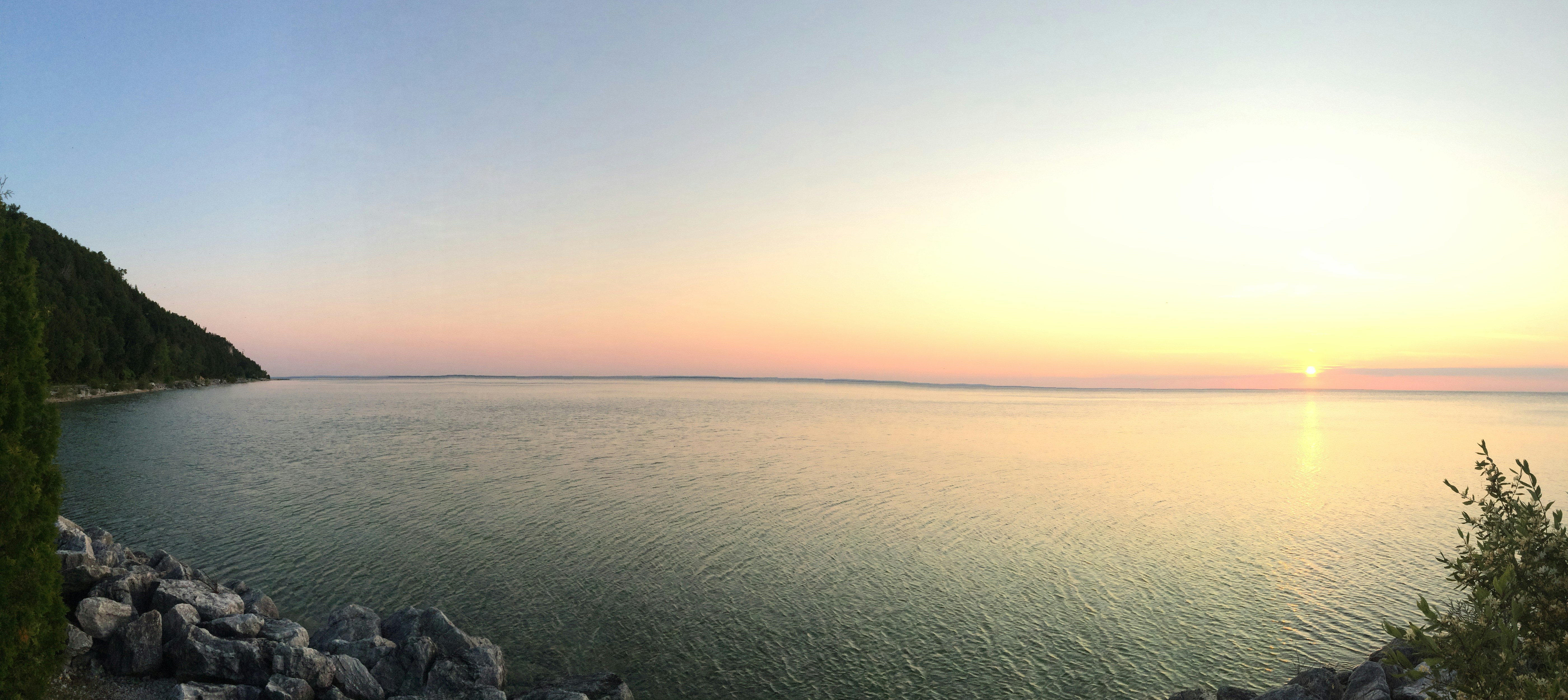 Panoramic view of a tranquil lake at sunset, with gentle waves reflecting soft hues of pink and orange. Lush greenery frames the scene.