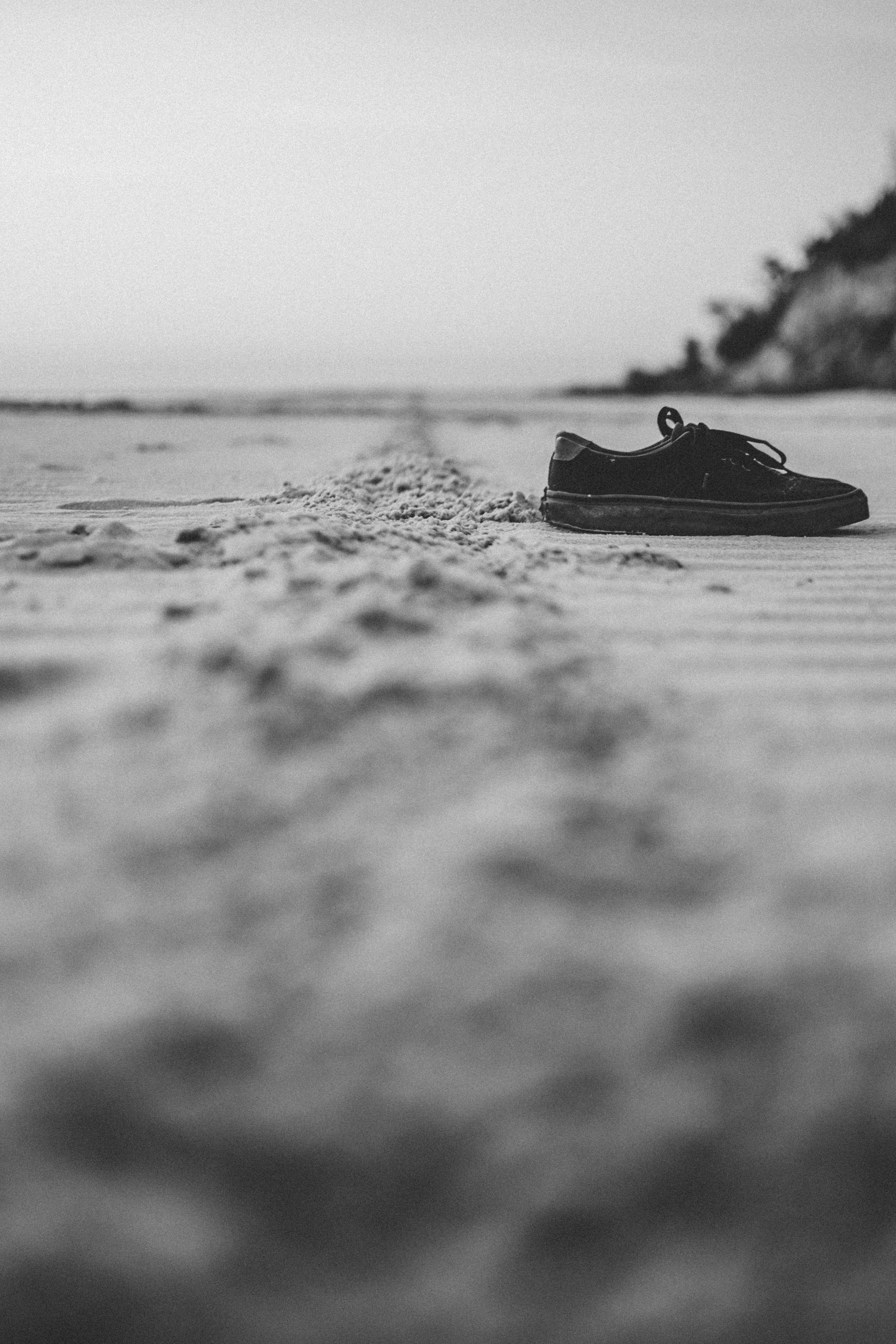 A lone black shoe rests on a sandy beach, with soft waves lapping in the background. The monochrome tones enhance the serene atmosphere.