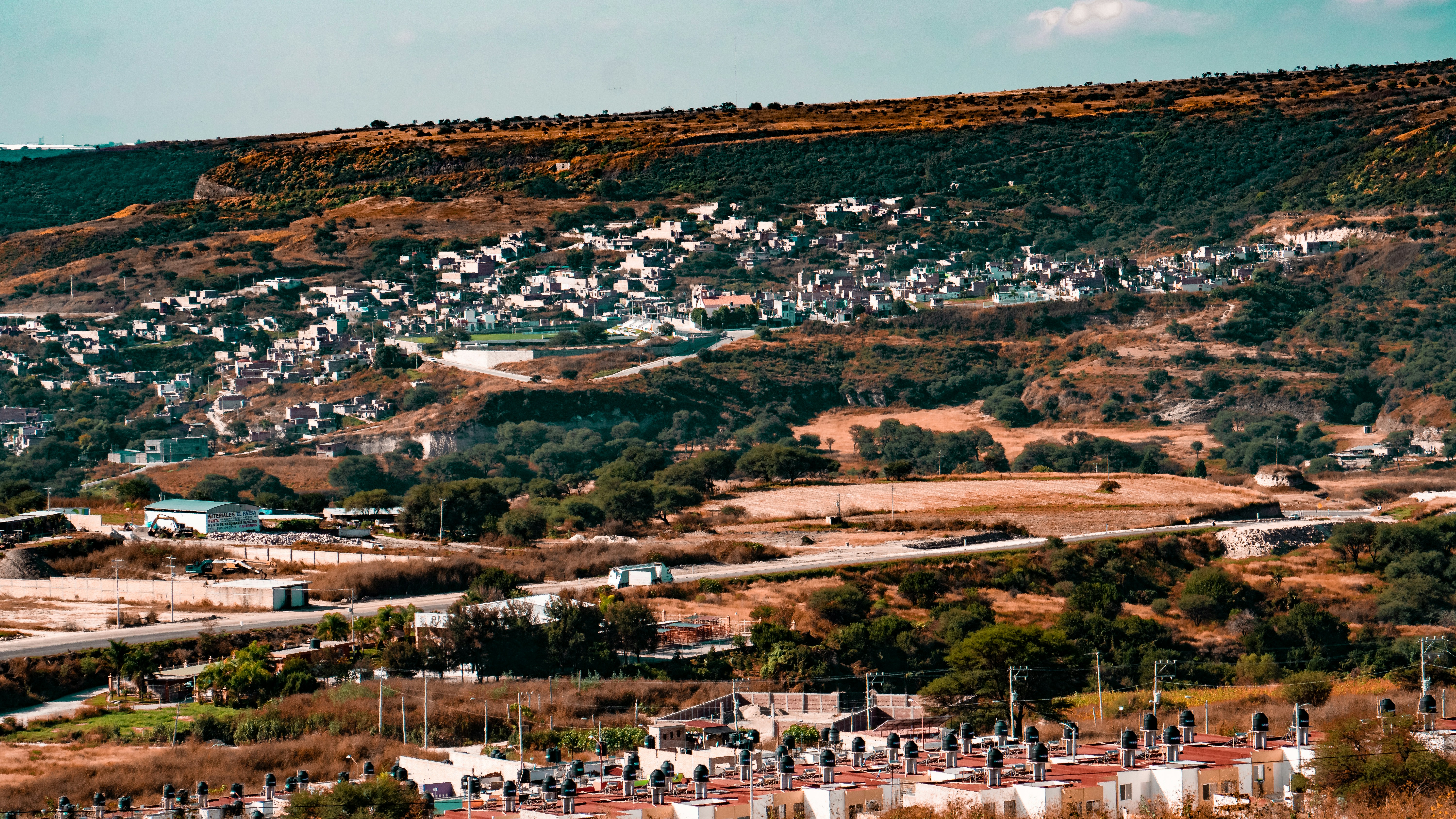 Panoramic view of a hillside town with scattered white buildings and surrounding greenery under a blue sky.