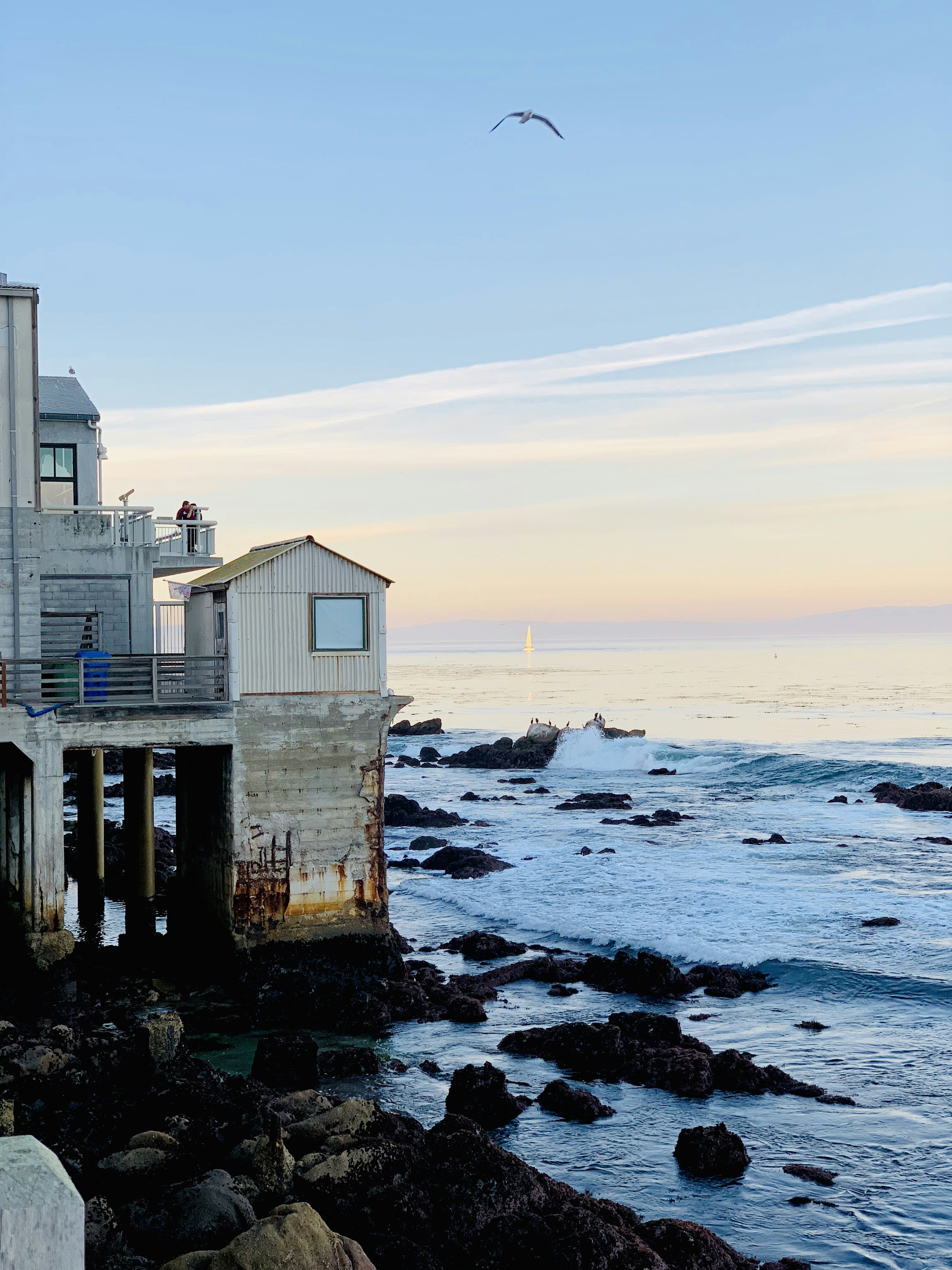 A coastal structure perched above rocky shores, with waves crashing below and a distant sailboat on the horizon.