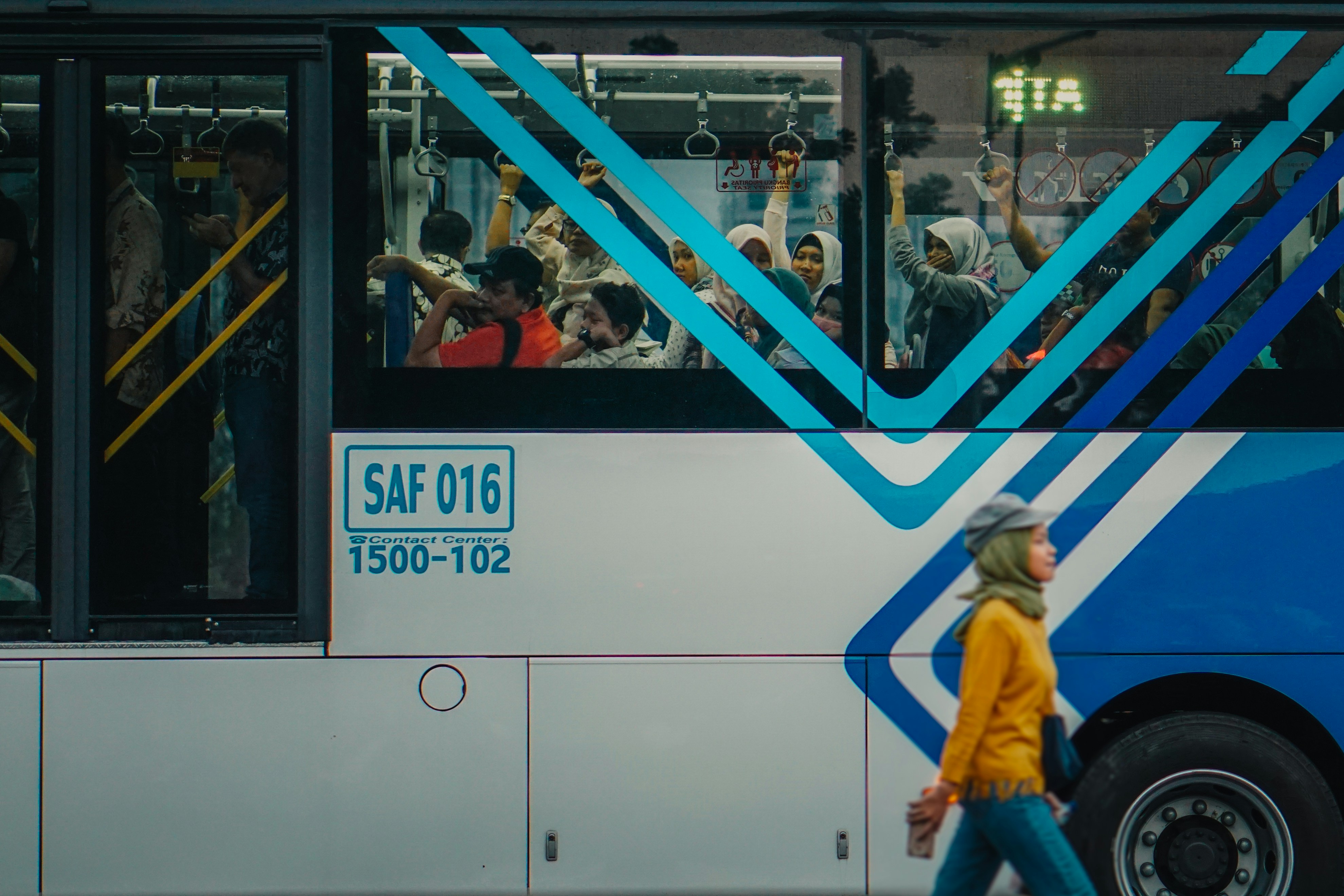 Commuters packed inside a city bus as a person walks past in vibrant attire.