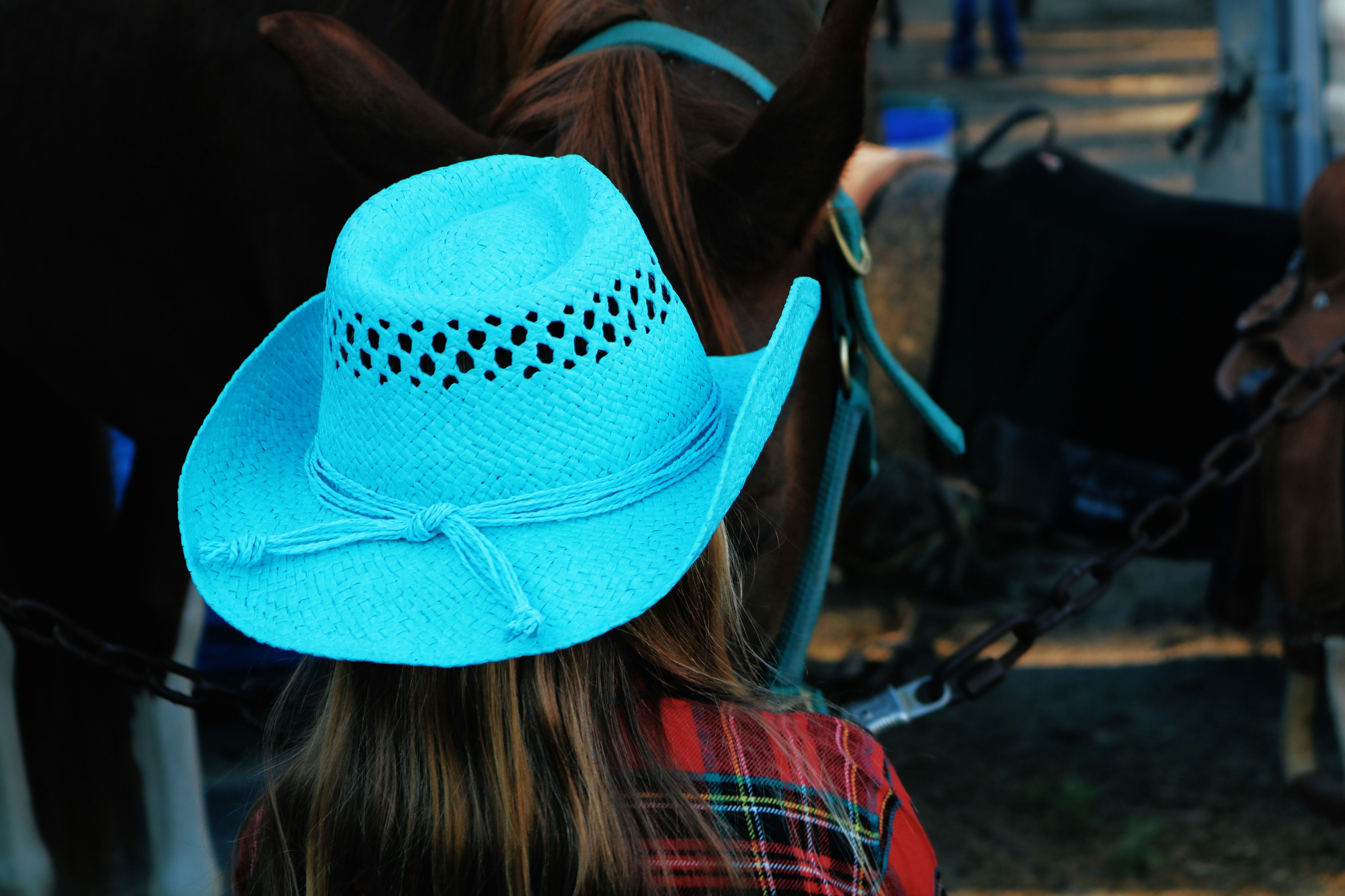 Child wearing a vibrant aqua cowboy hat gazes at horses in the background, capturing the essence of rural life and rodeo culture.