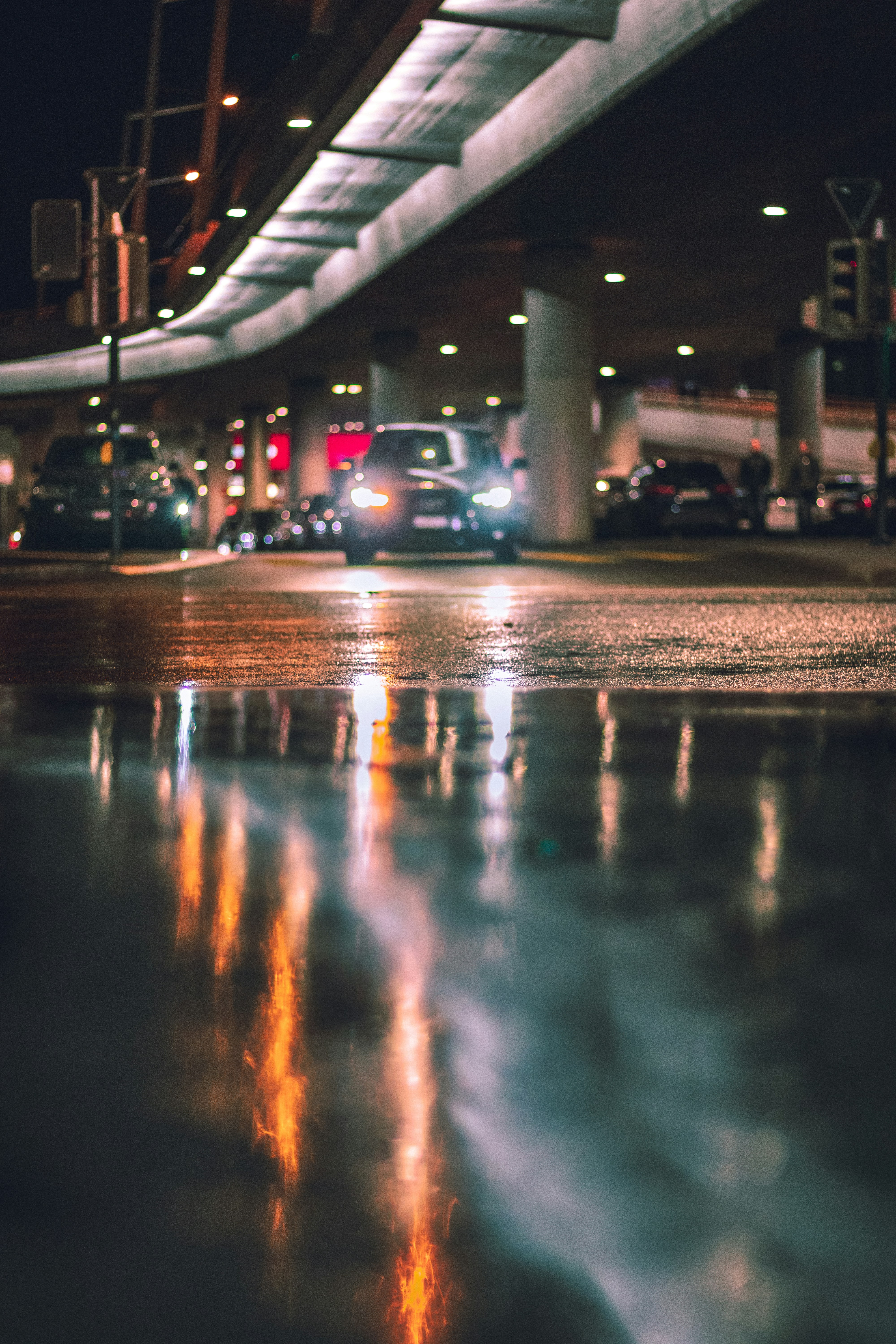 Rain-soaked pavement reflecting car lights and urban structures under a highway overpass.