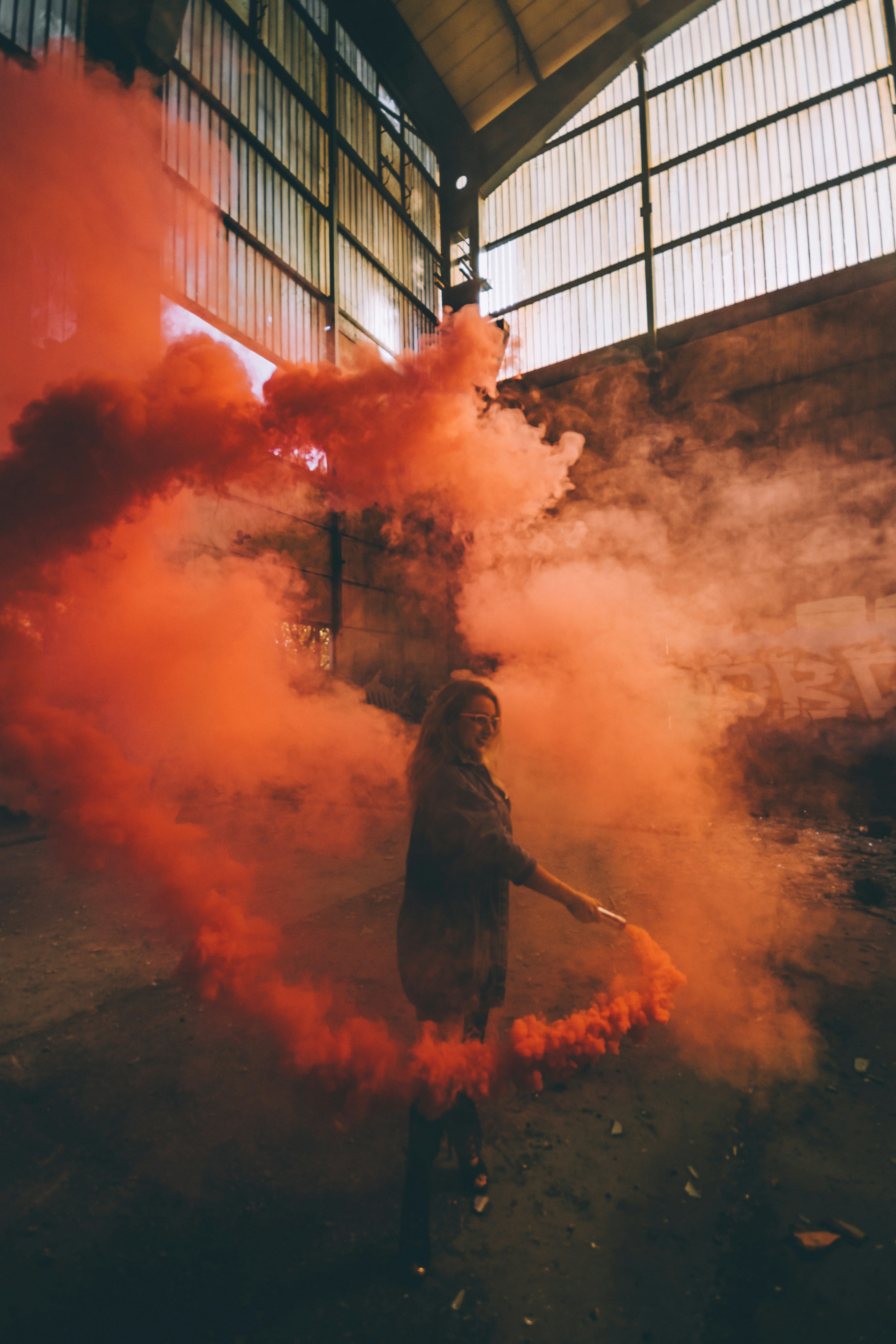 A person twirls with a smoke flare, enveloped in vibrant orange clouds within an industrial space. The interplay of light and shadow enhances the dynamic atmosphere.