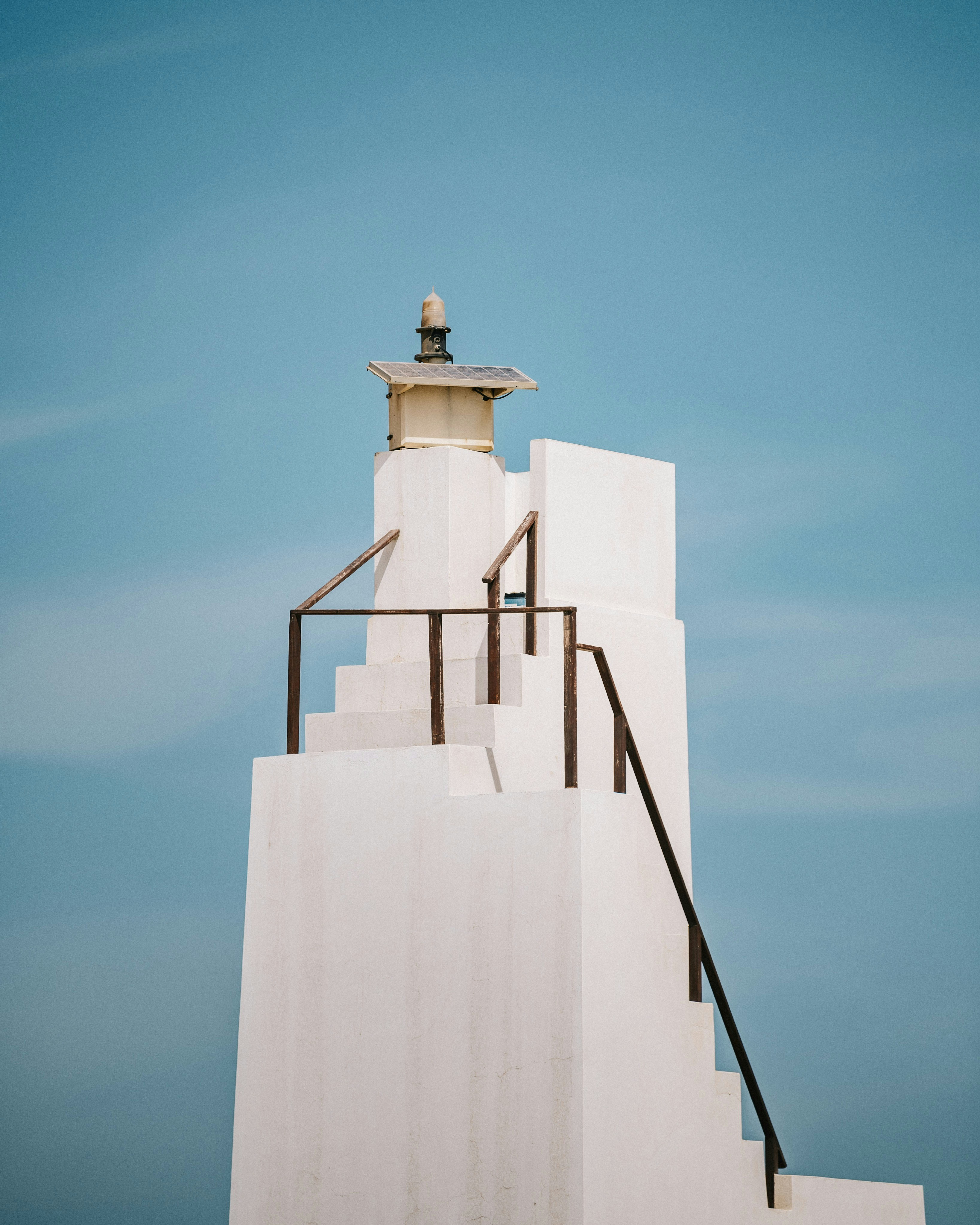 A minimalist white lighthouse with a solar panel, featuring a staircase leading to the light atop against a clear blue sky.