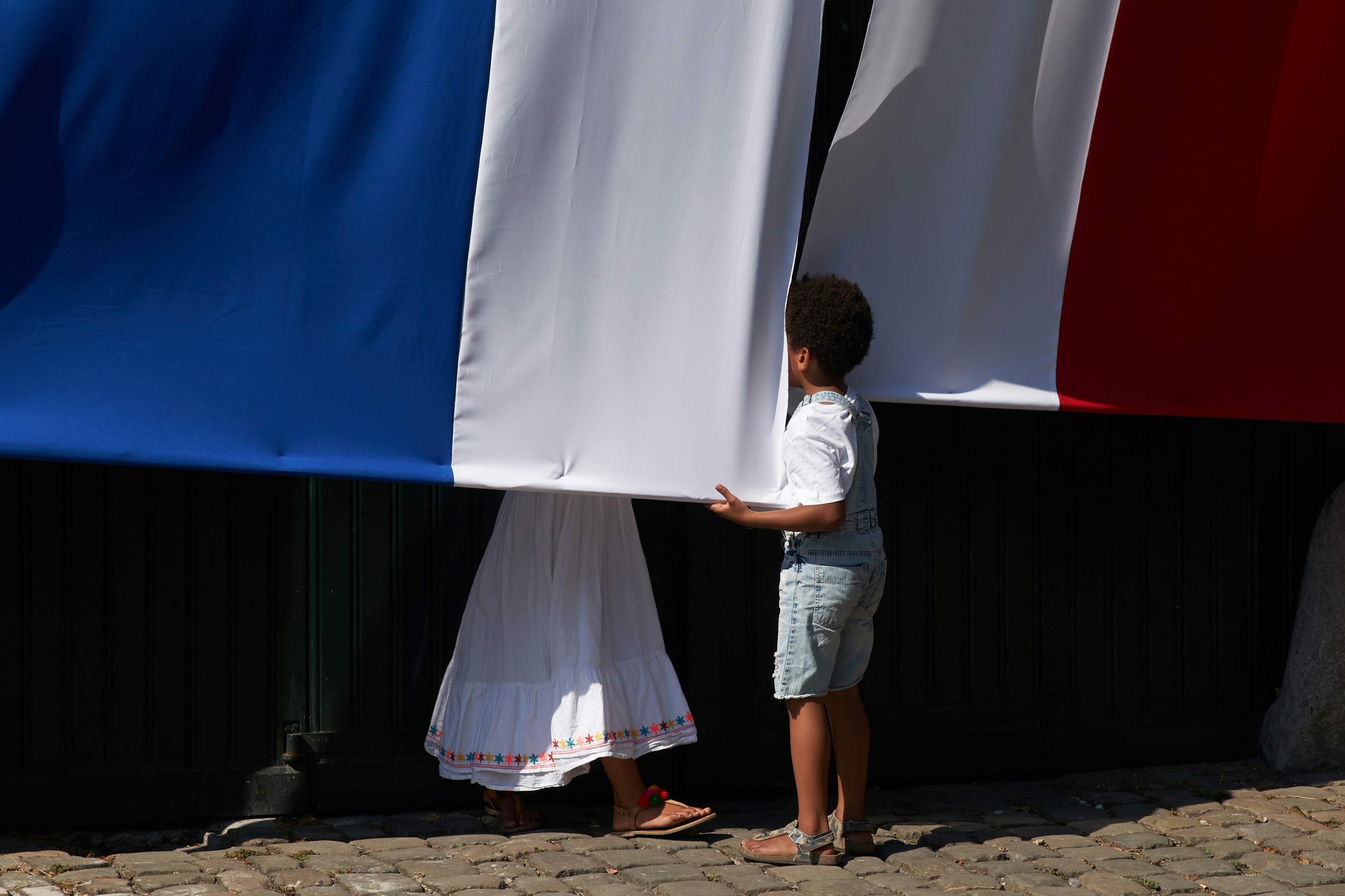 A child curiously peers behind a large French flag, while a figure in white stands partially concealed. The scene captures a moment of intrigue and cultural connection.