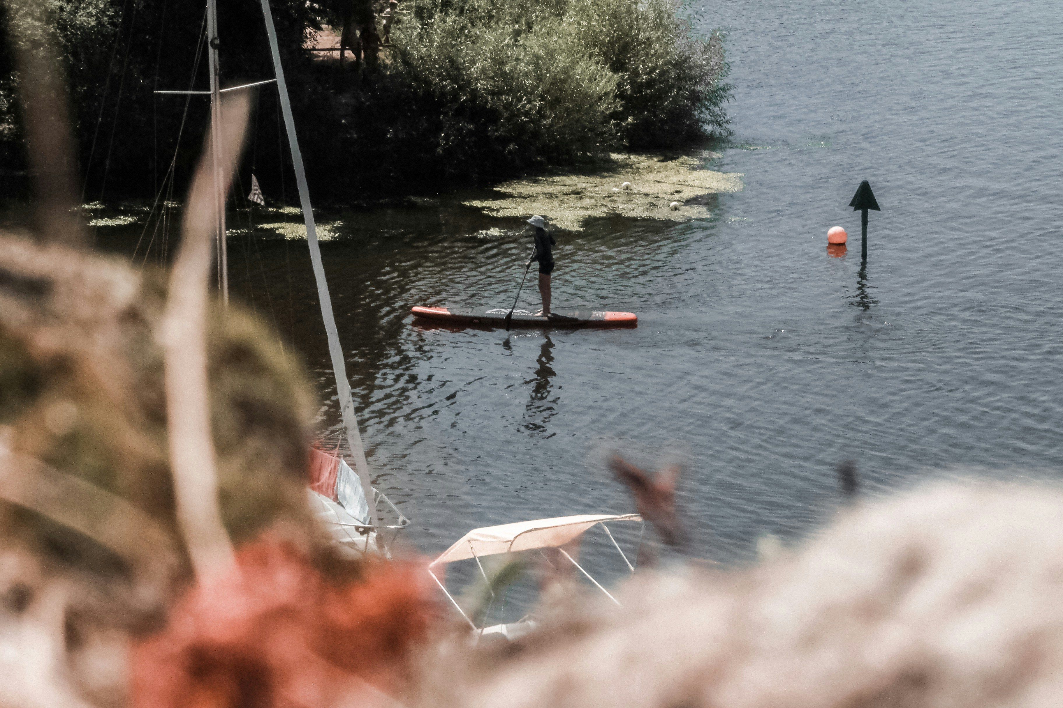 Color photograph of a lone paddler standing on a red stand-up paddleboard on a calm lake, foregrounded by blurred vegetation. A buoy marks a quiet focal point in the water.