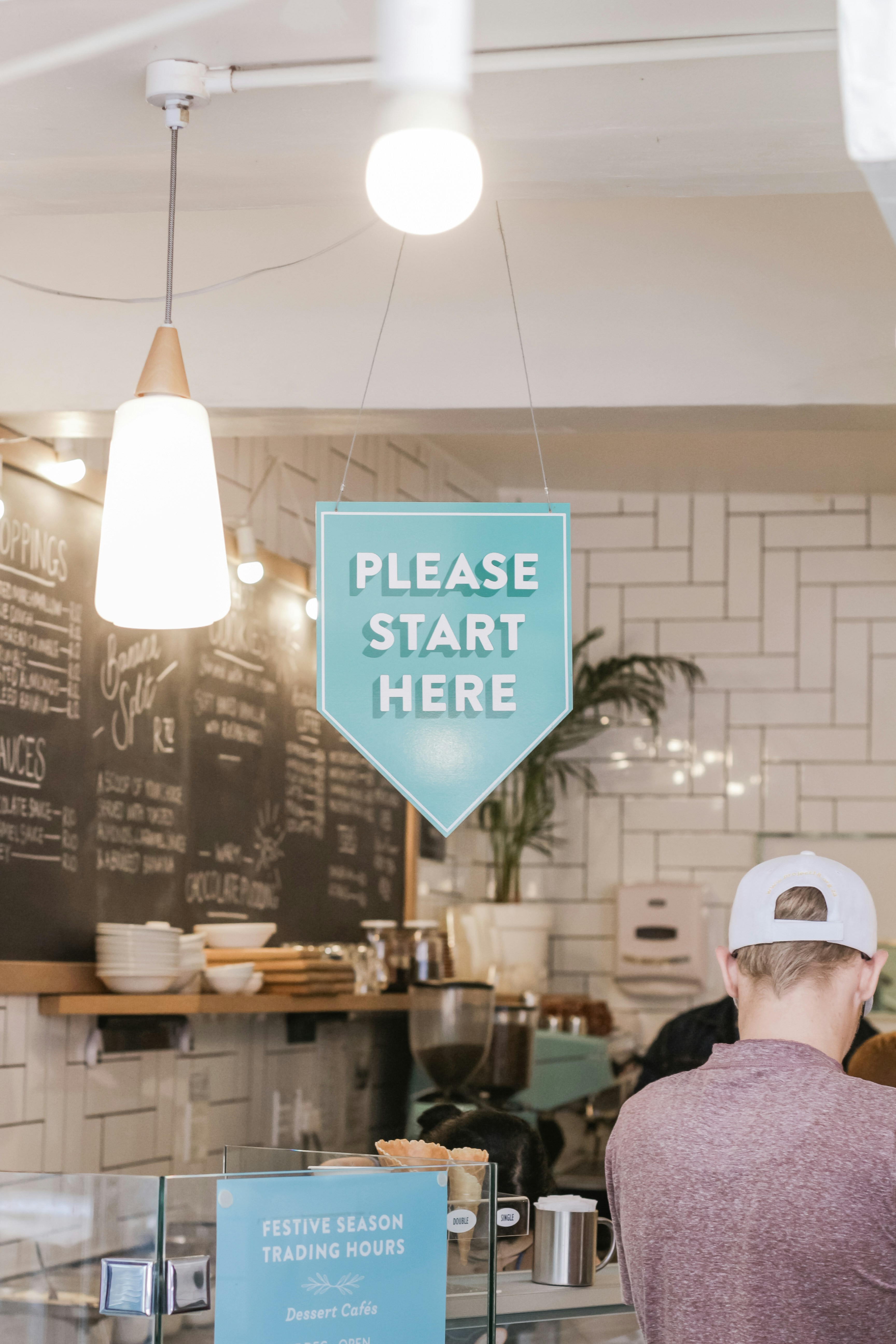 A teal sign reading 'PLEASE START HERE' hangs in a cozy café, surrounded by a vibrant atmosphere of menu boards and warm lighting.