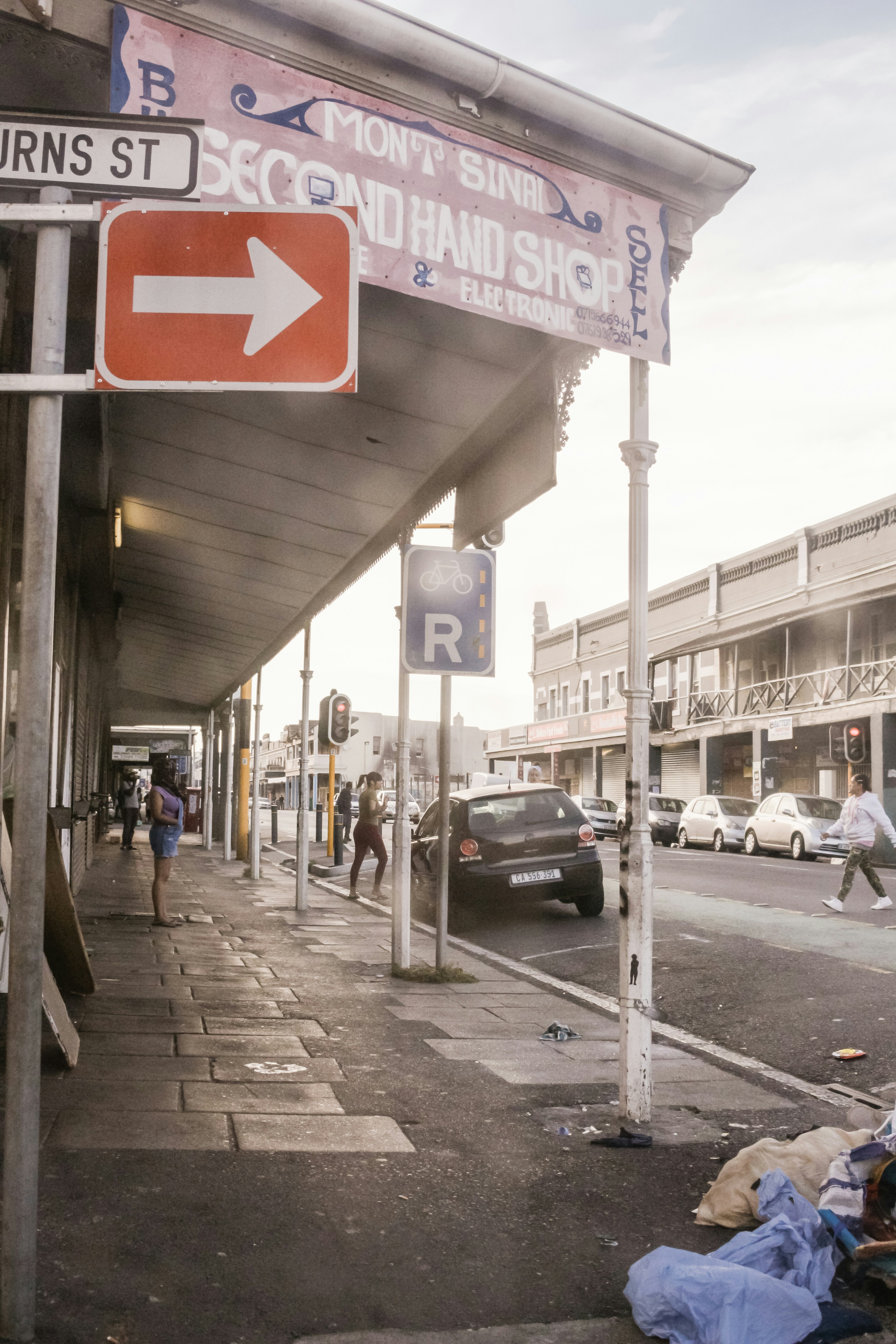 A bustling street scene featuring a second-hand shop sign and pedestrians navigating the sidewalk. The atmosphere captures the essence of urban life.
