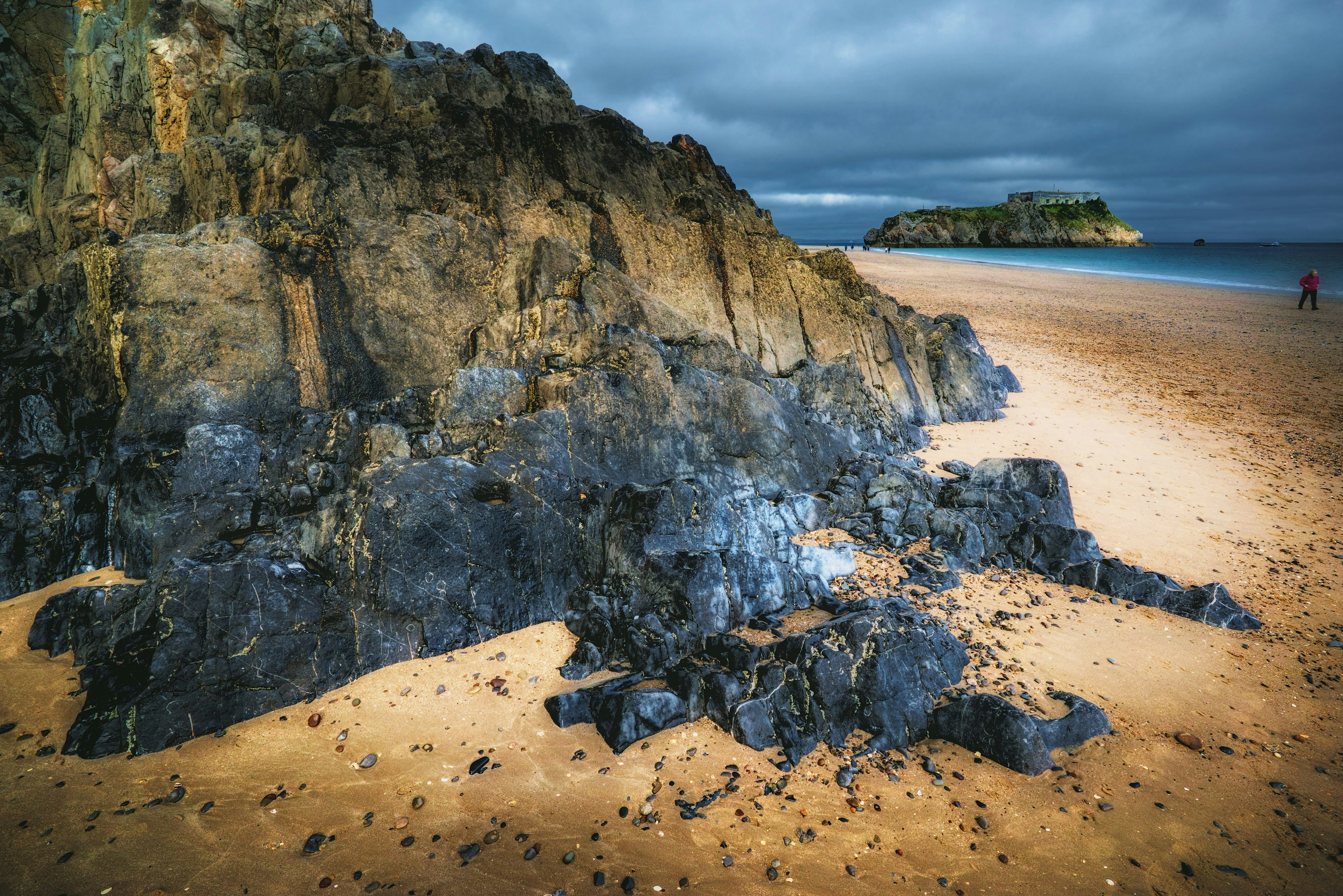 Jagged rocks with intersecting patterns on a sandy beach lead to St. Catherine's Island under a dramatic cloudy sky.