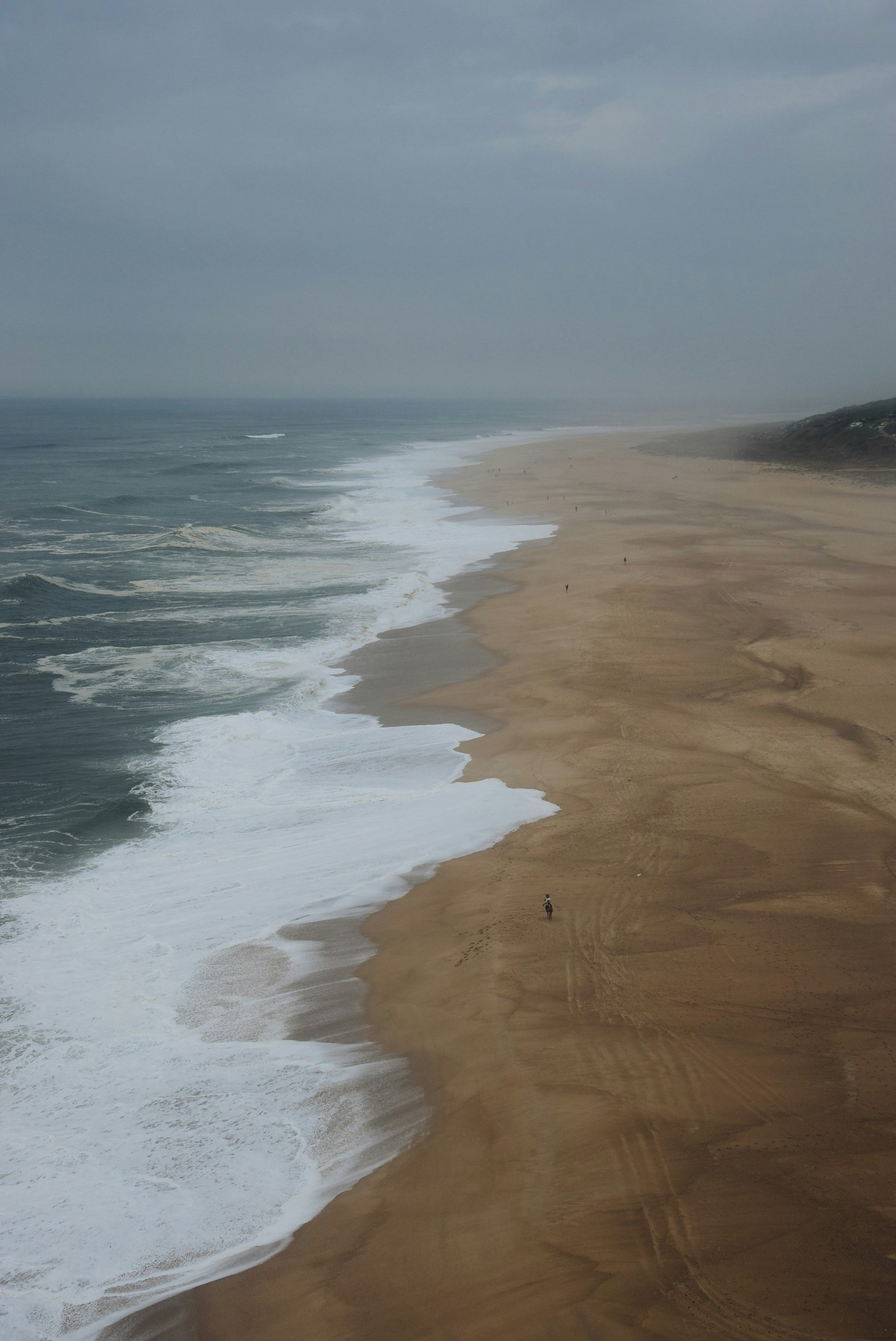 Waves gently lap against a vast sandy beach under a moody sky, with distant figures strolling along the shore. The scene evokes a sense of tranquility and solitude.