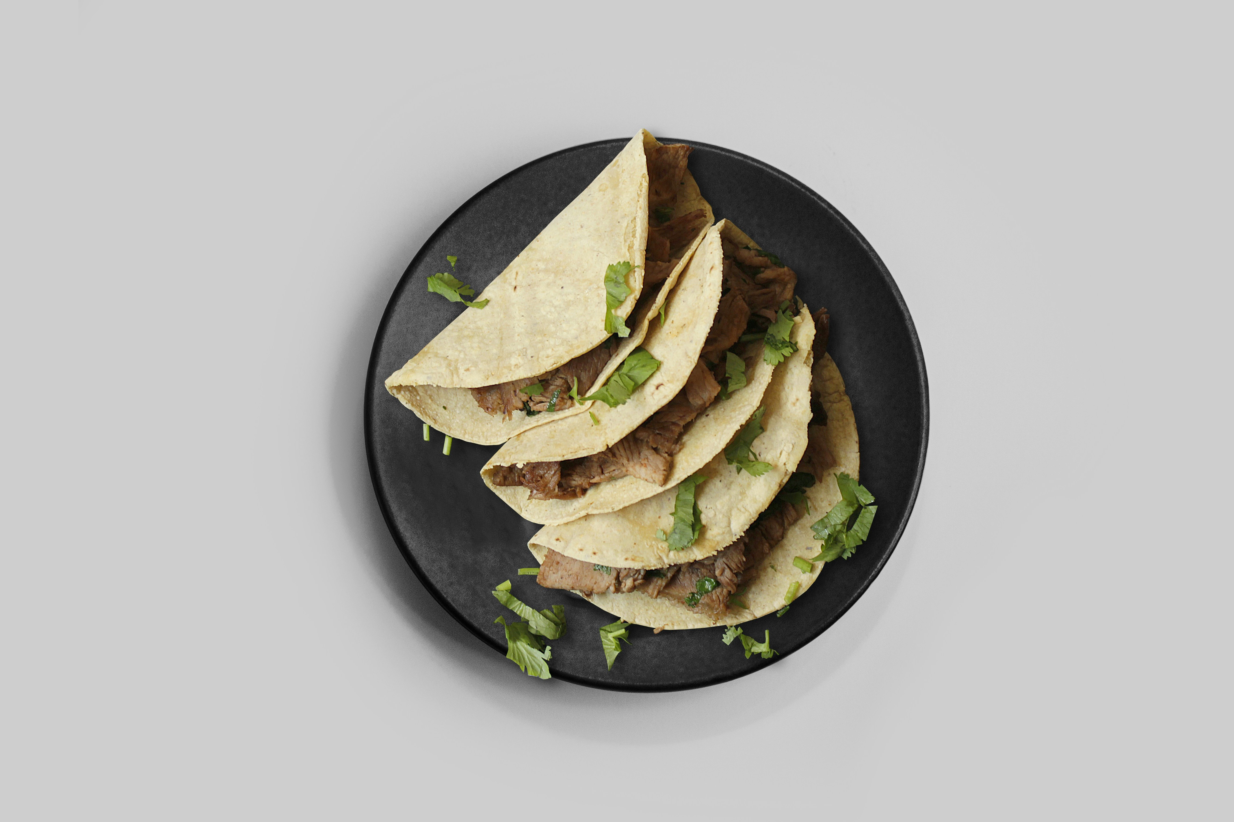 Four tacos arranged on a black plate with fresh cilantro garnish.