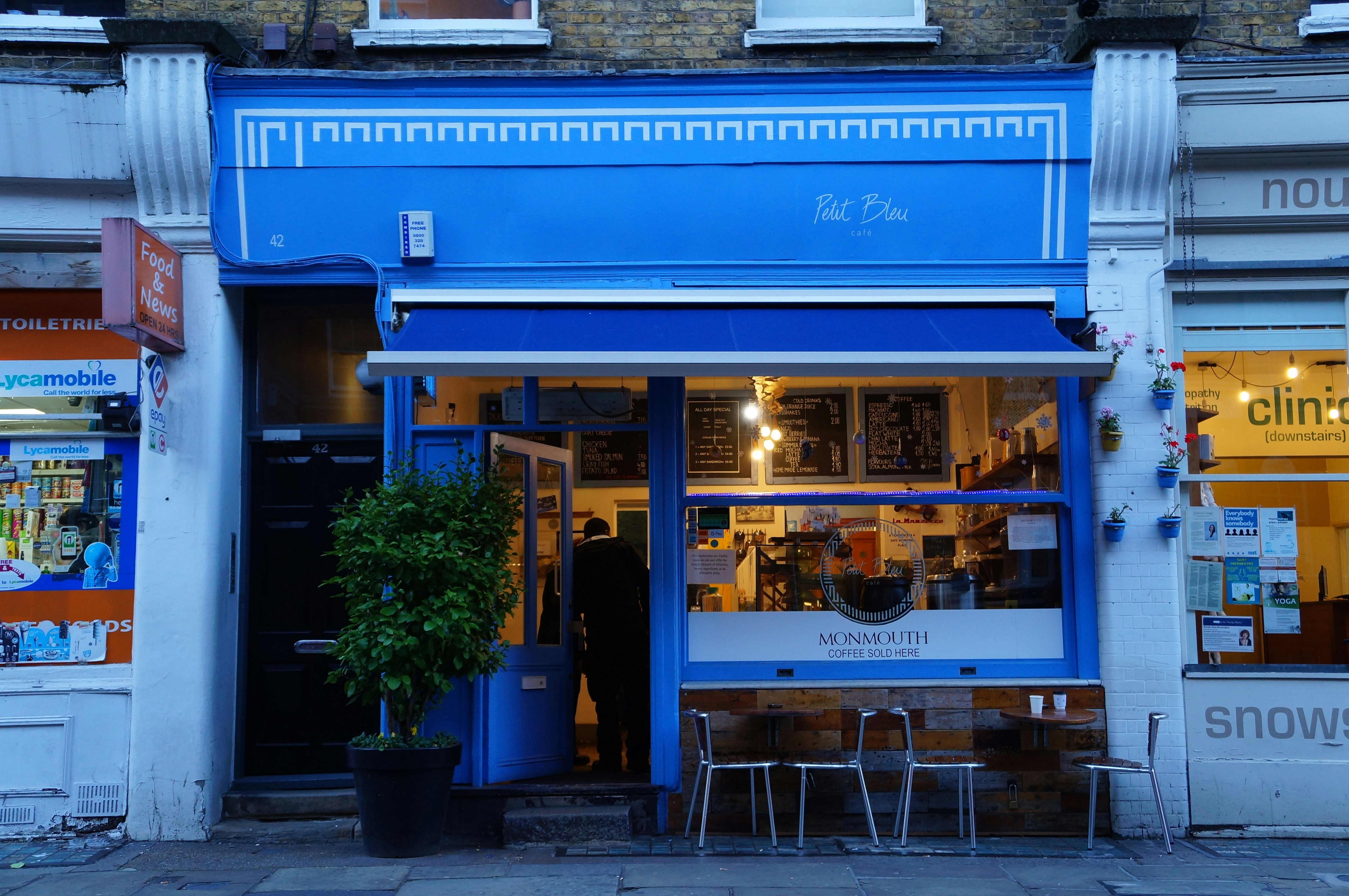 Blue-fronted cafe with open doors and warm interior lighting on a bustling street.