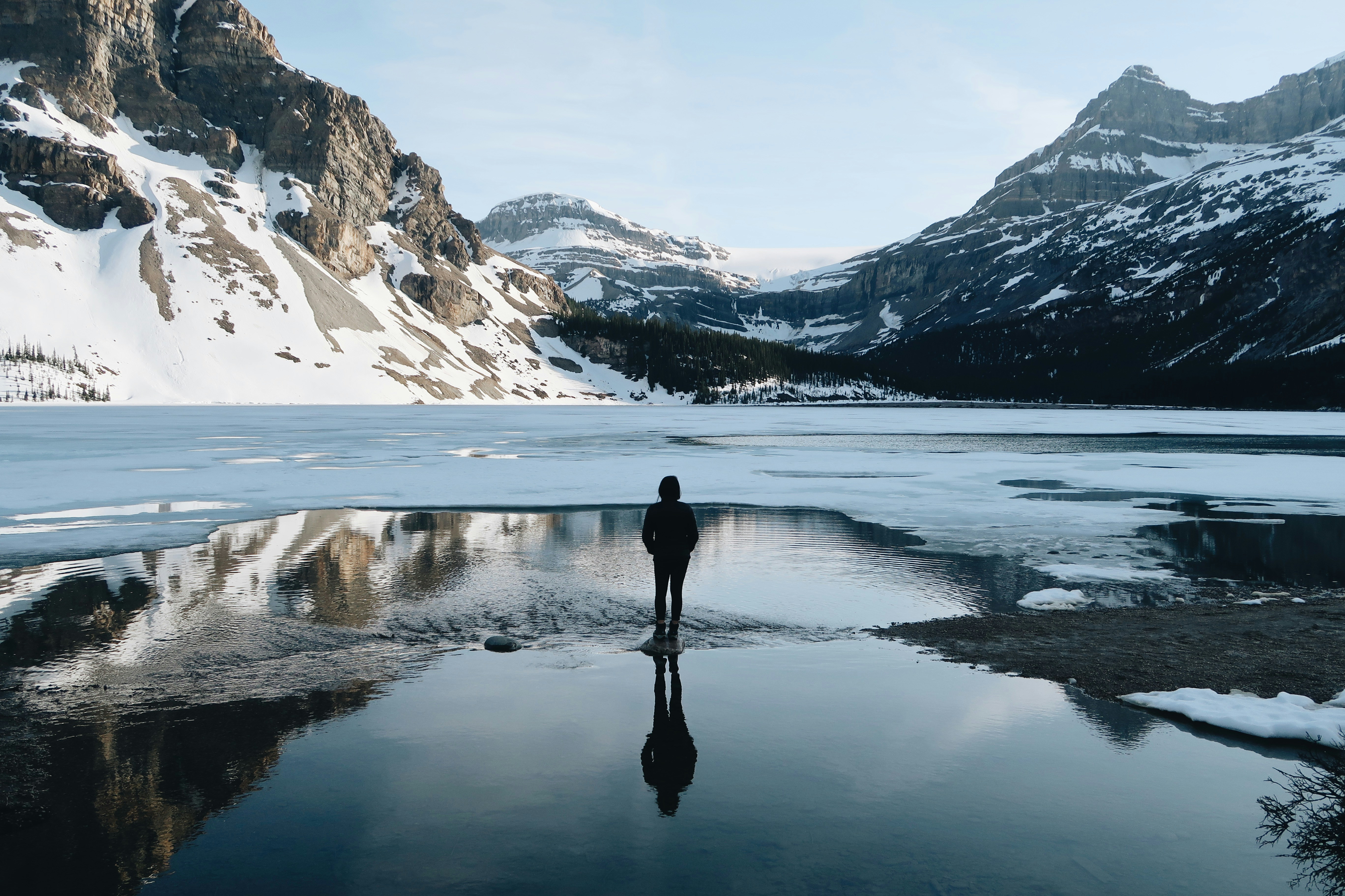 A solitary figure stands on the edge of a partially frozen lake, surrounded by majestic snow-capped mountains reflecting in the water.