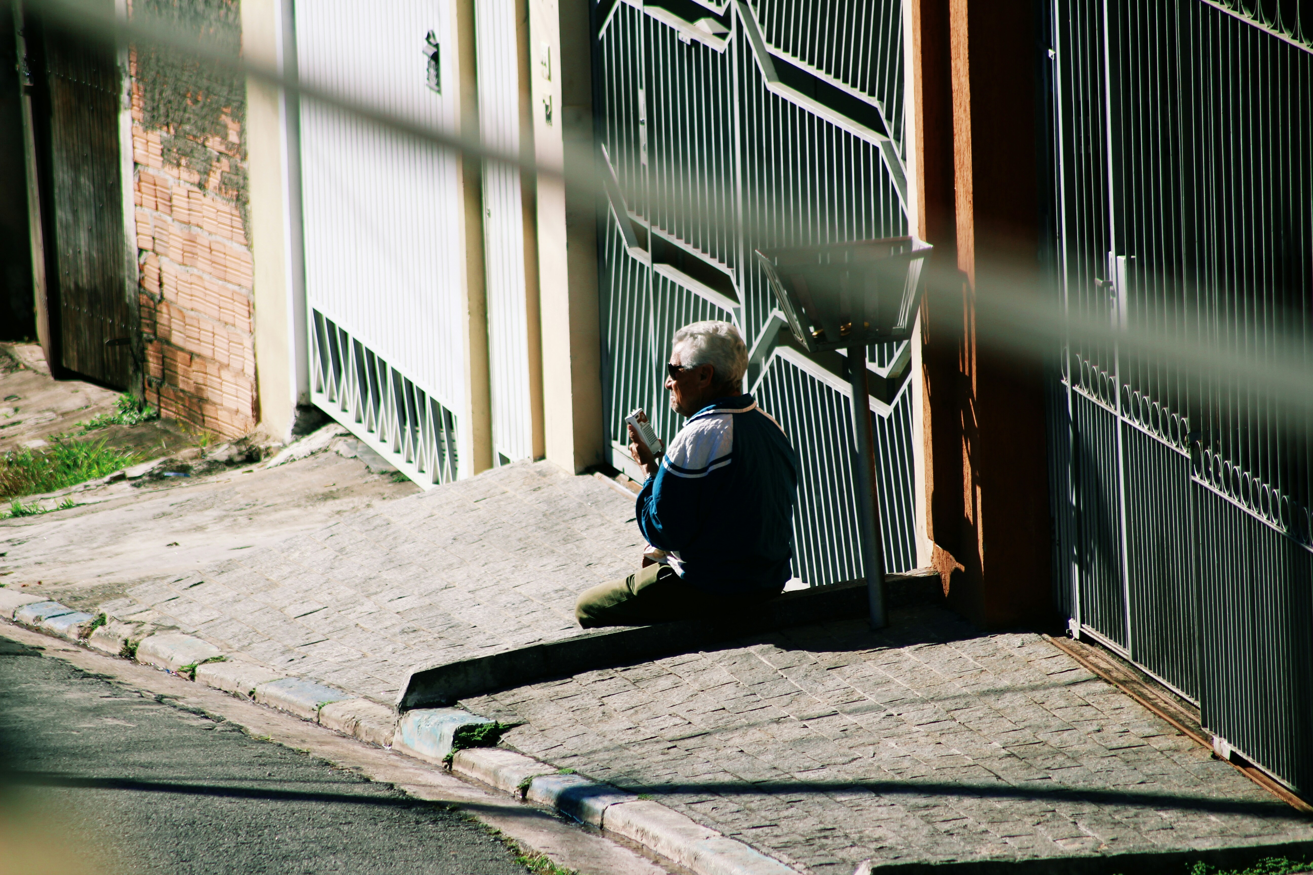 An elderly man sits on the pavement, absorbed in thought against a backdrop of a gated entrance. The scene captures a quiet moment in urban life.