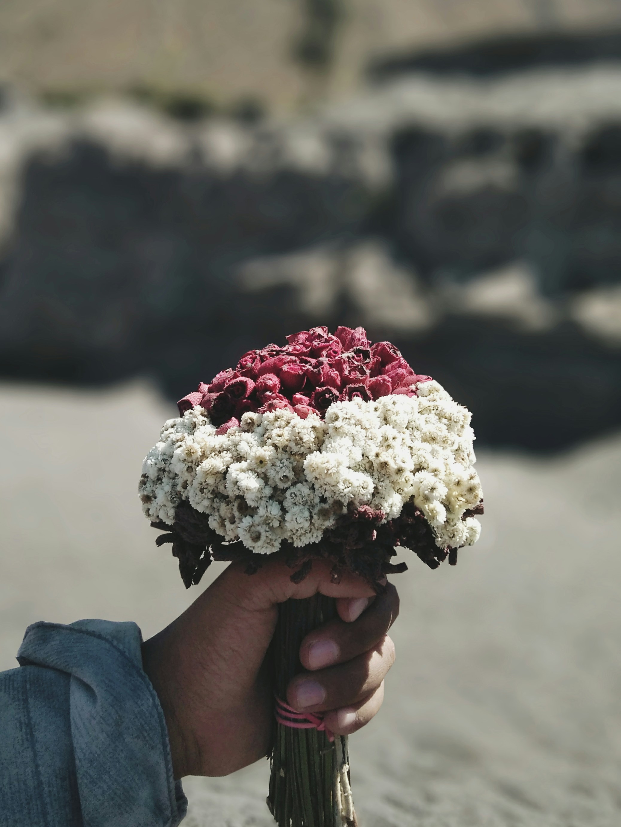 Close-up of a hand gripping a small bouquet of dried crimson and ivory flowers, set against a softly blurred stone backdrop.