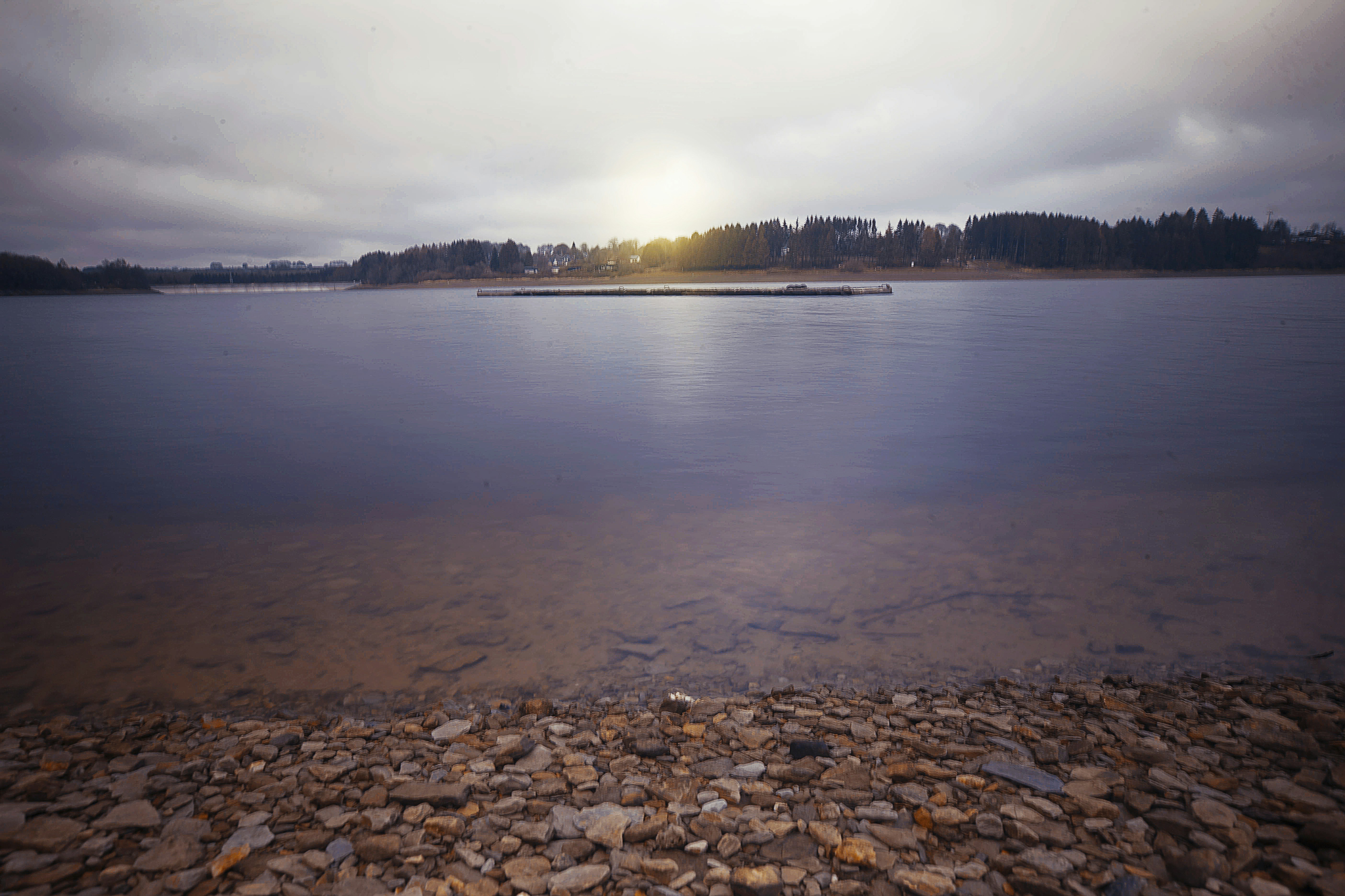 Sunrise over calm lake waters with a rocky shoreline under a cloudy sky.