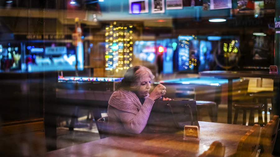 An older woman sits alone at a café table, leaning over a cup amid colorful reflections and blurred city lights outside the window.
