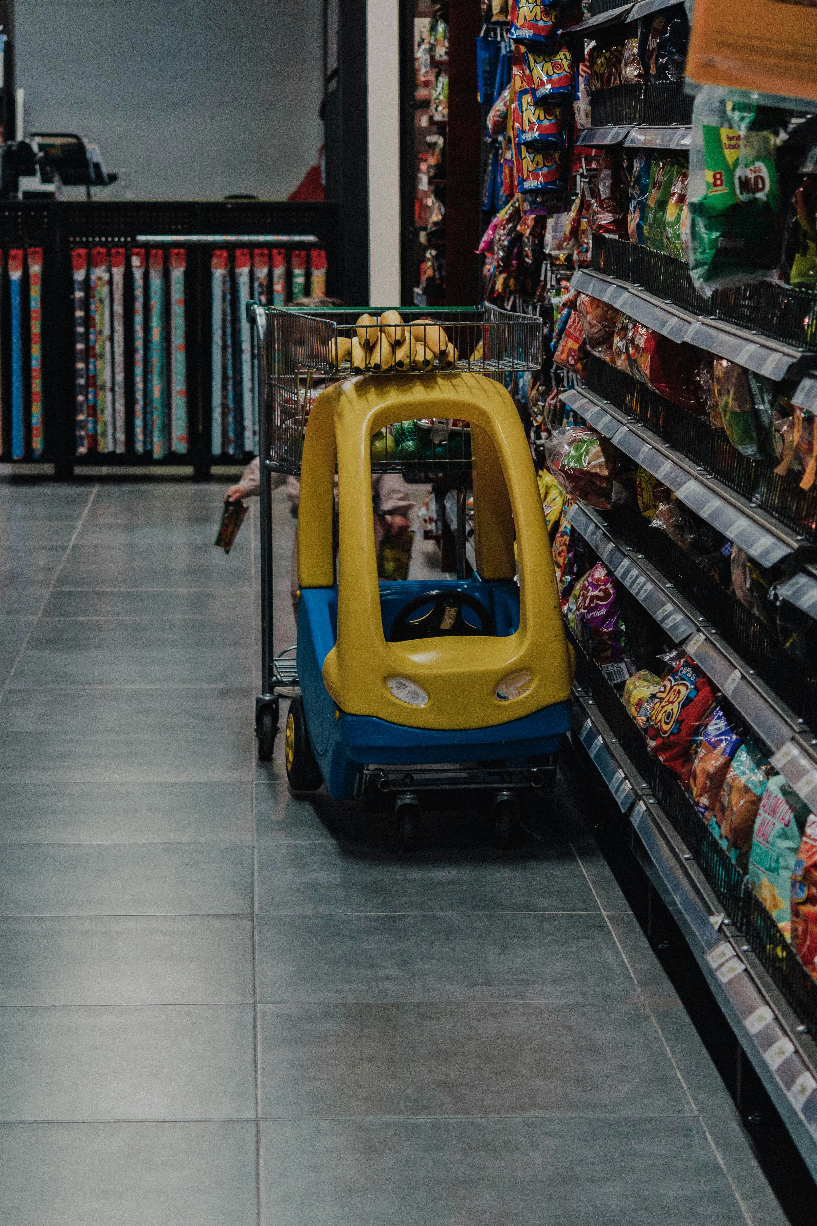 A colorful toy car cart filled with bananas parked beside an aisle of snacks in a grocery store.