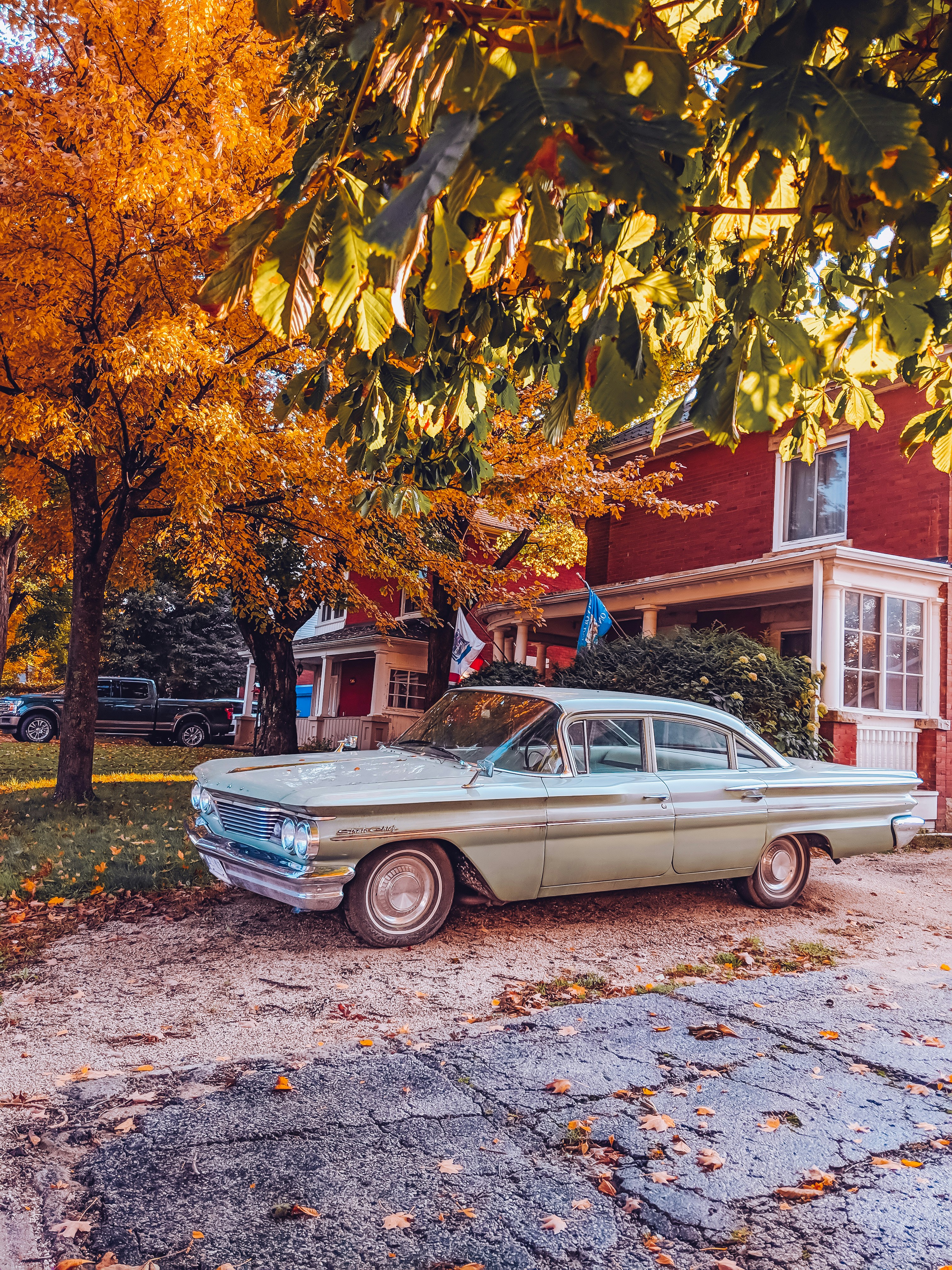 Classic green car parked beside vibrant autumn trees in front of a historic house. The scene captures the essence of fall with colorful foliage.