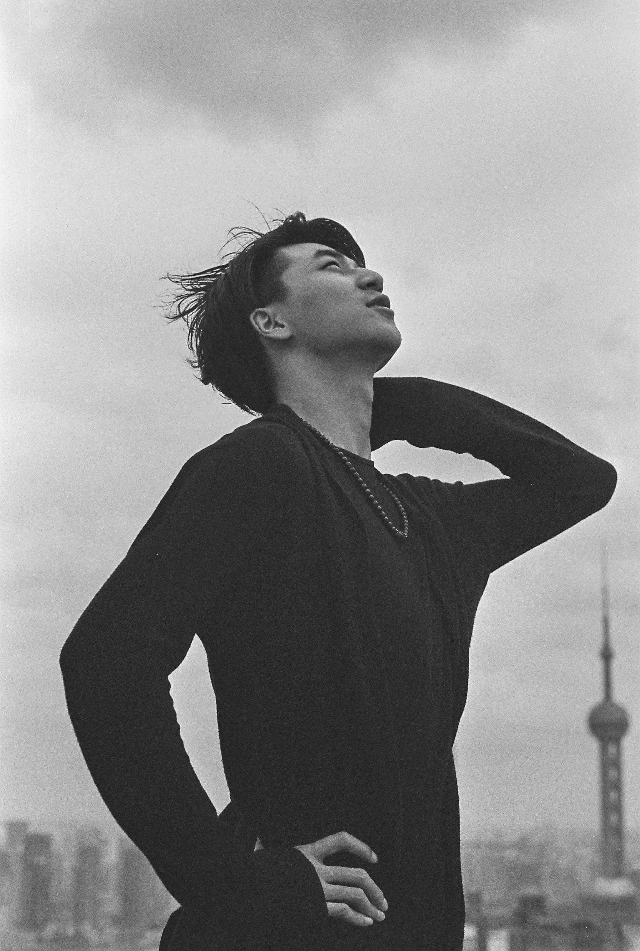 A young man stands confidently against a cloudy skyline, his head tilted upwards, exuding a sense of contemplation and poise.