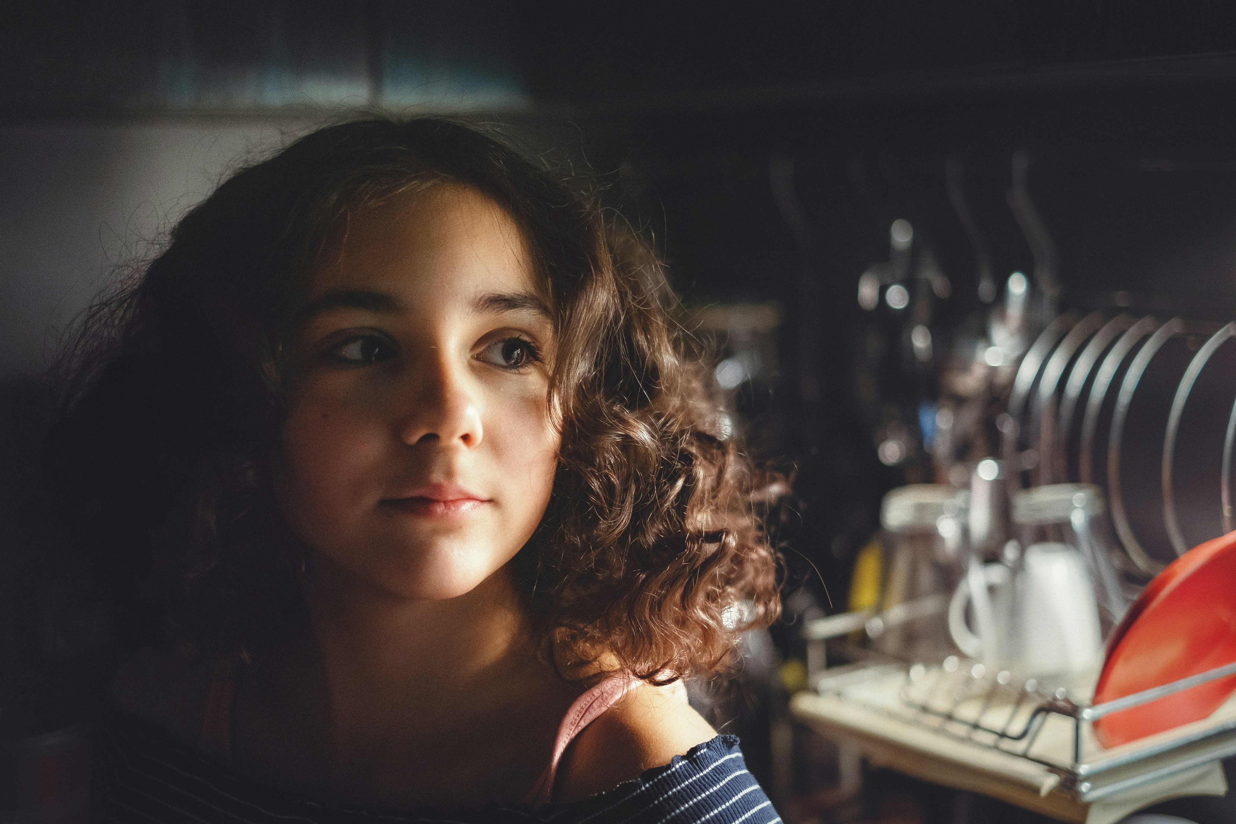 Young girl with curly hair gazes thoughtfully, illuminated by soft light amidst kitchen utensils and dishware.