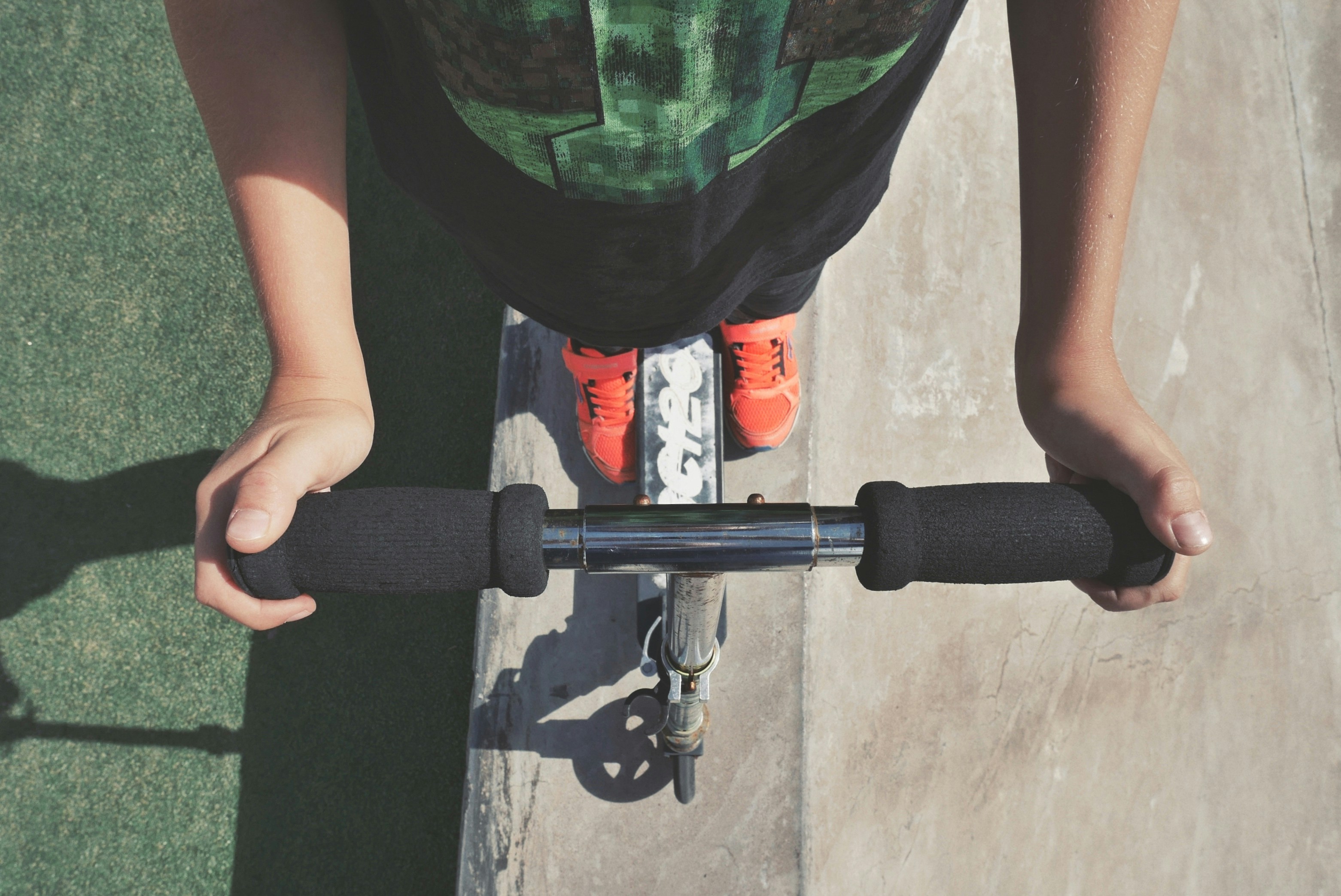 Child's hands gripping scooter handlebars with vibrant sneakers visible, set against a textured surface.
