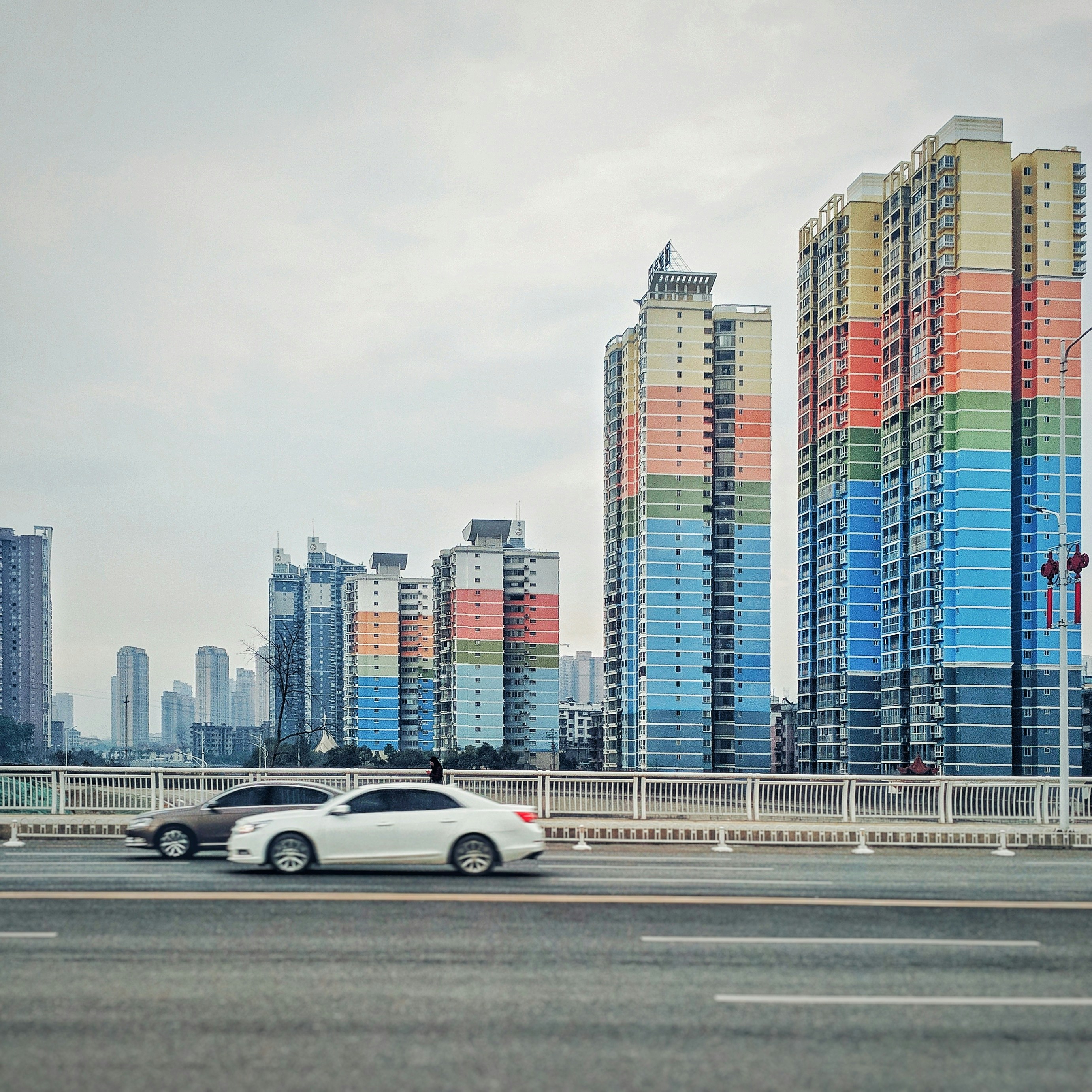 Rainbow-colored highrises rise beside a busy expressway. Blurred cars streak along the road, emphasizing motion against the skyline.