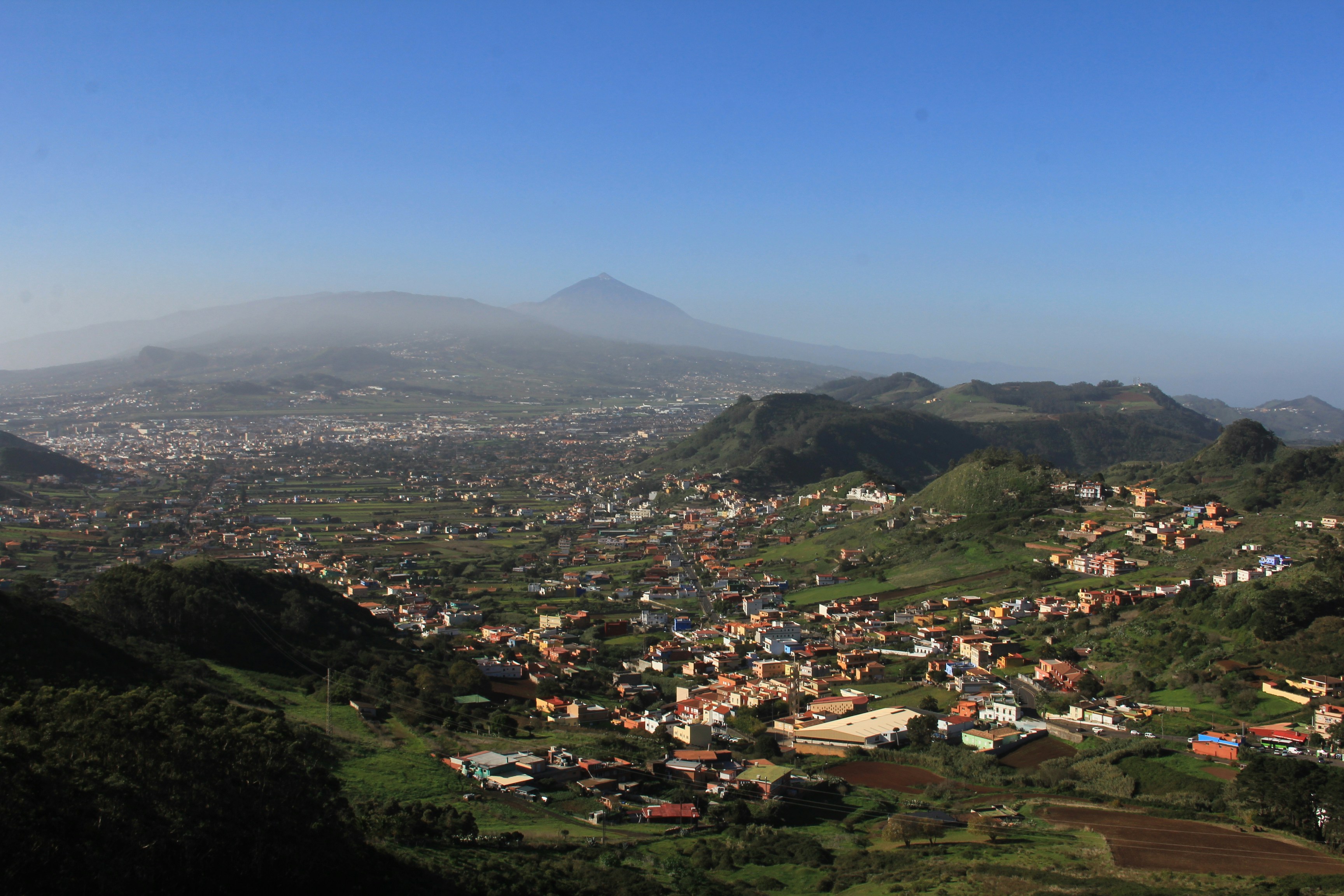 Vast landscape showcasing a patchwork of colorful homes nestled among rolling hills, with a distant volcano looming under a clear blue sky.