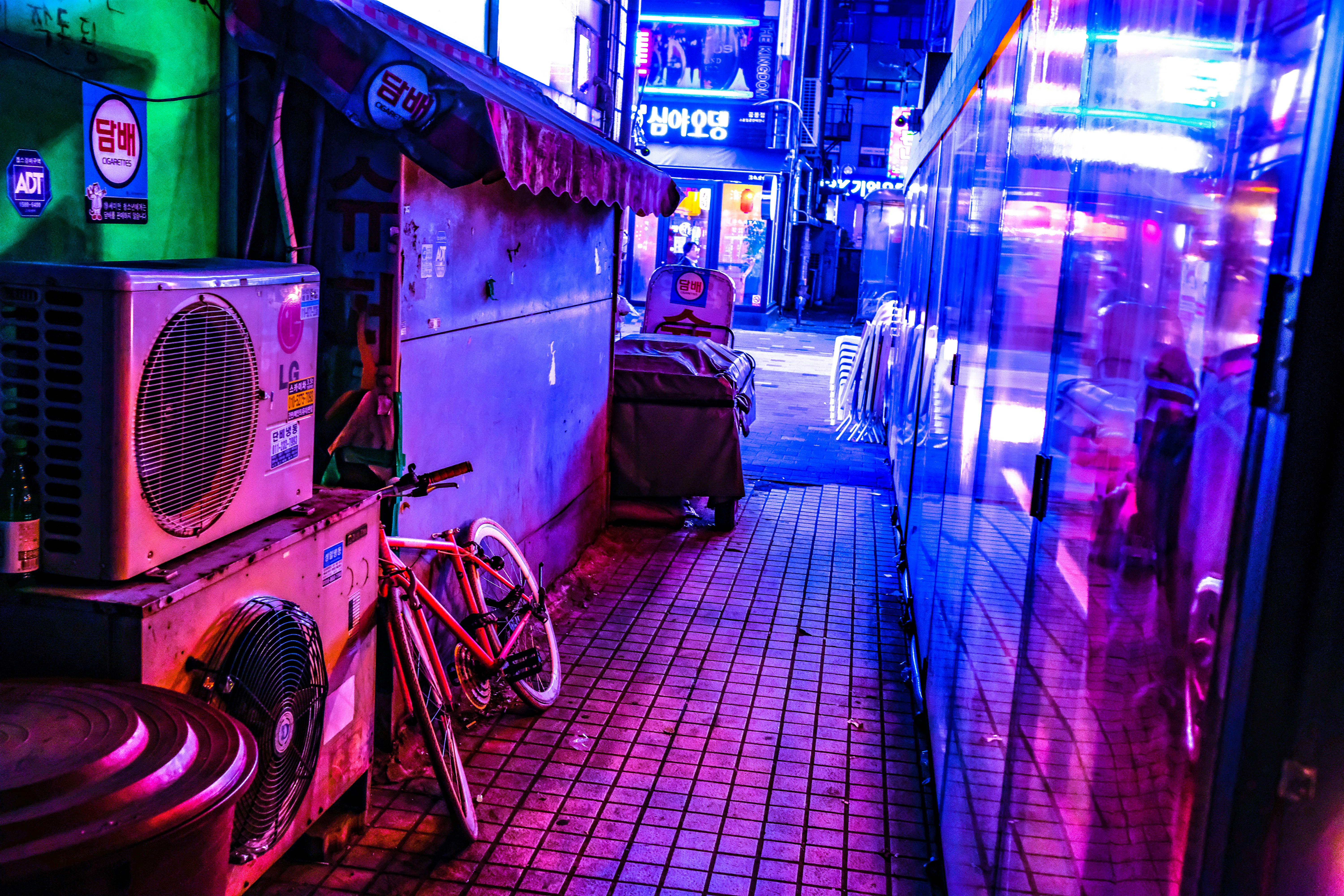 Vibrant alleyway illuminated by neon lights, featuring a bicycle and urban elements. The scene captures the essence of nightlife in a bustling city.
