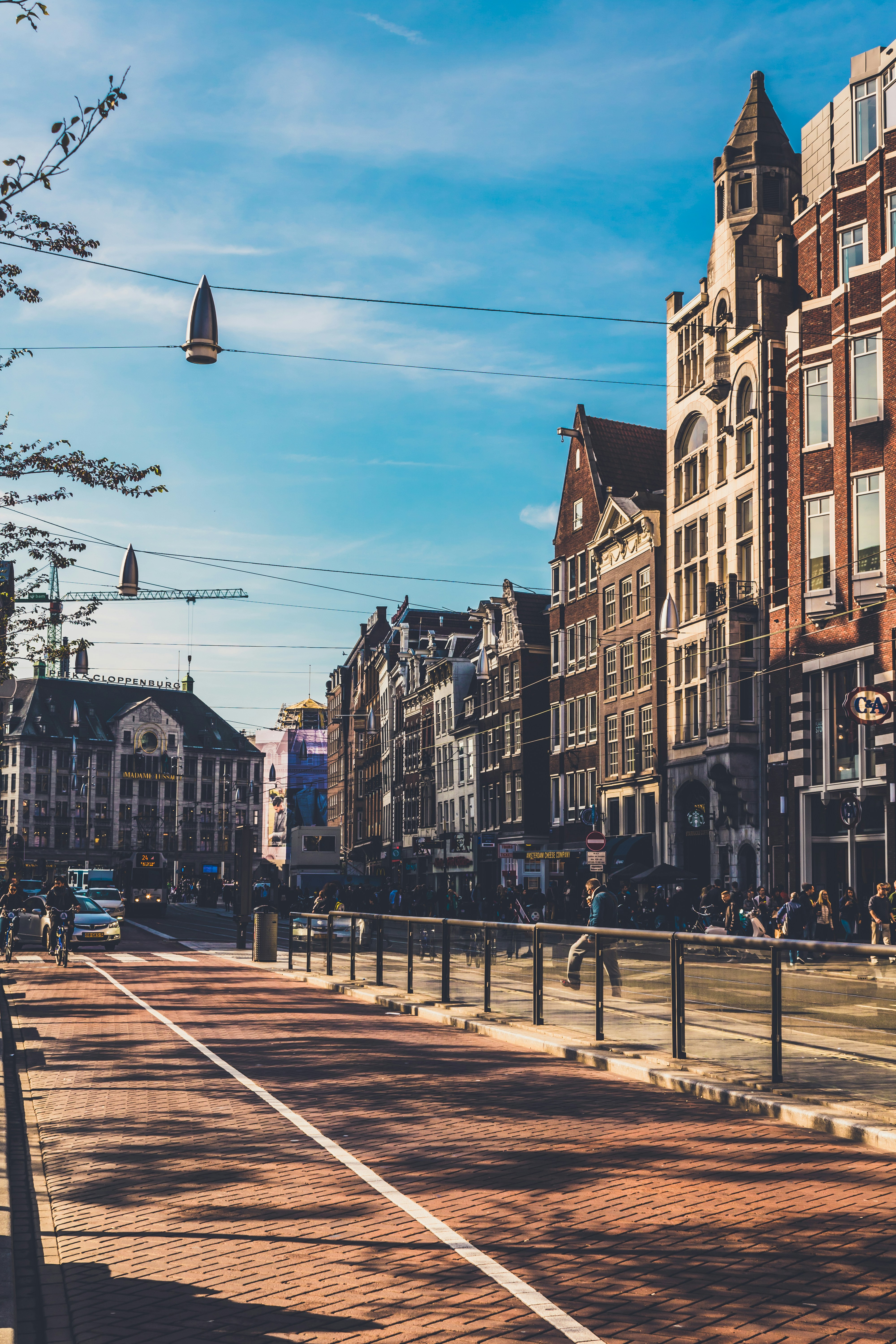 Historic buildings line a vibrant street under a clear blue sky, with pedestrians enjoying the lively atmosphere. Street lamps and construction cranes add to the urban scene.