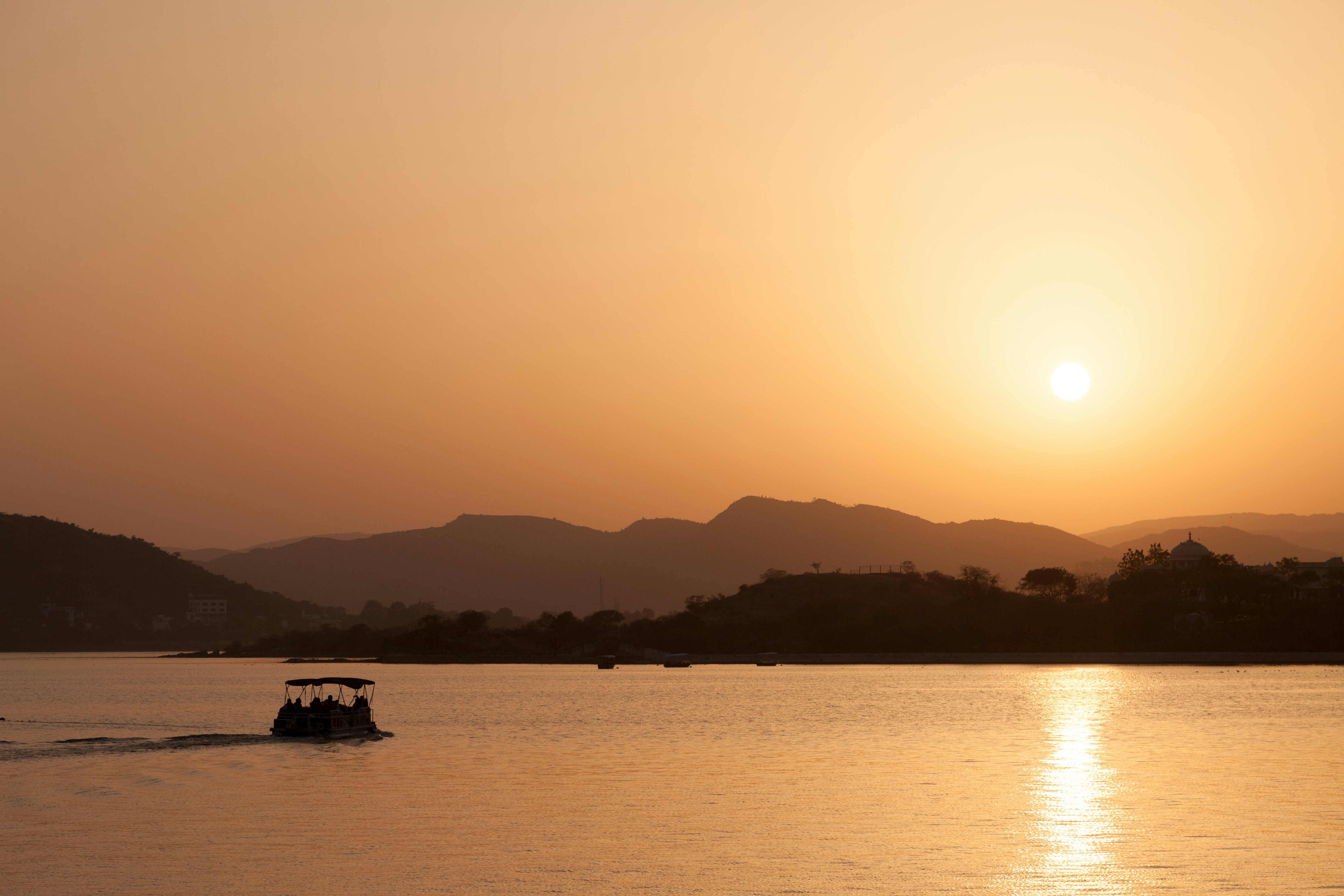 A boat glides across a tranquil lake at sunset, with silhouettes of distant mountains framing the scene. The sun casts a warm glow over the water's surface.