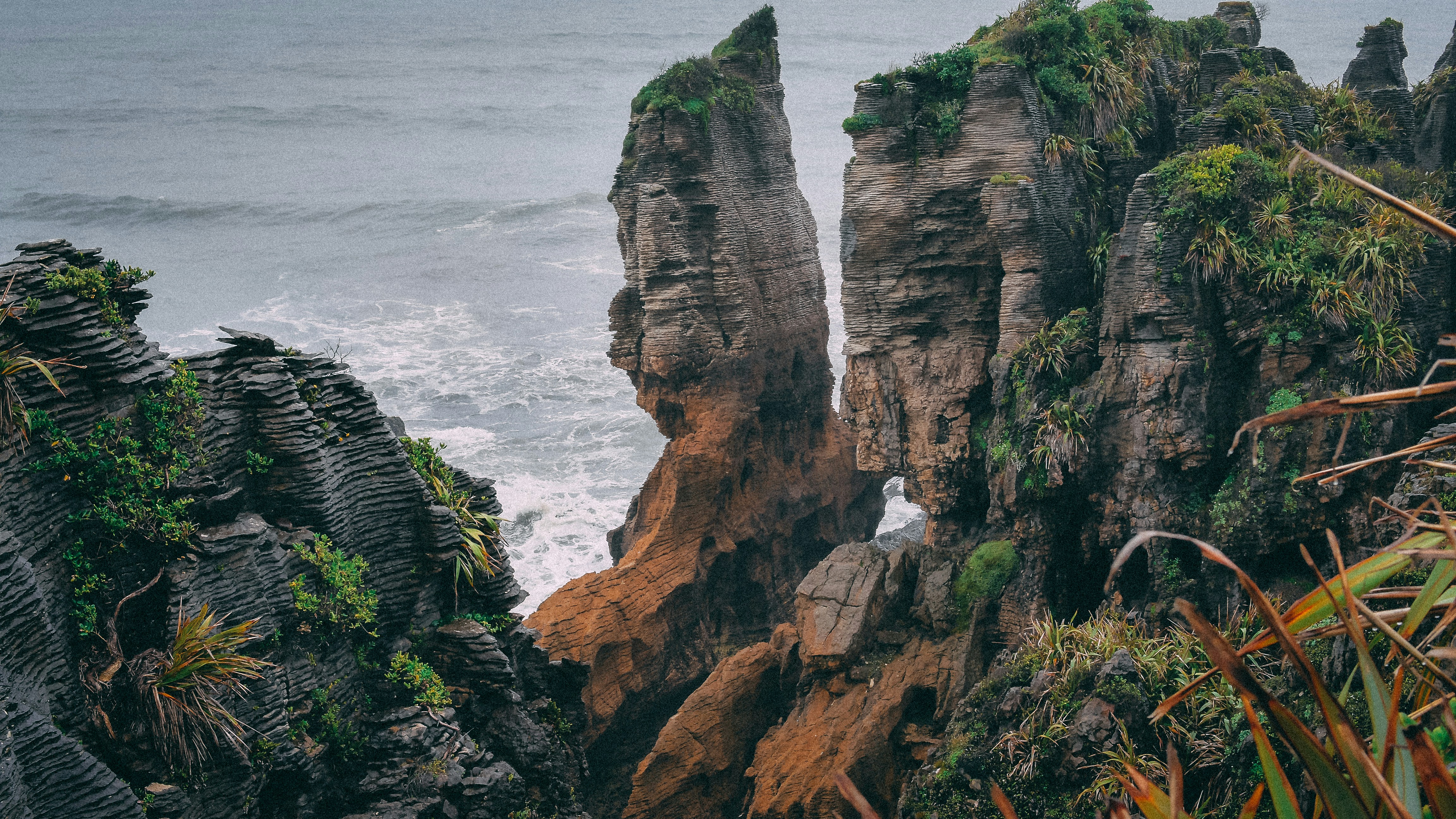 Rugged rock formations stand sentinel over crashing ocean waves, adorned with lush greenery. The scene captures the raw beauty of coastal erosion and resilience.
