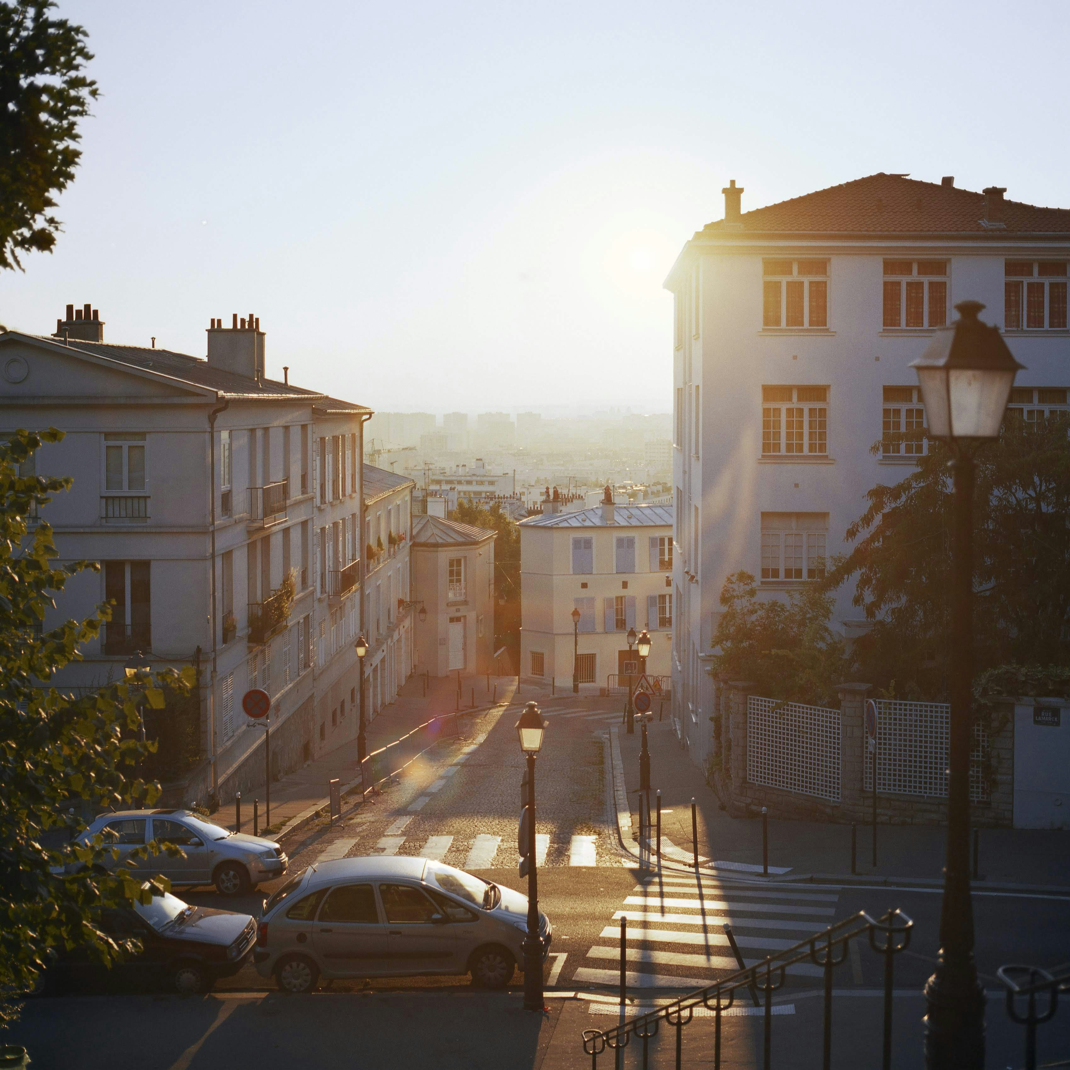 Sunlight bathes a quiet street lined with charming buildings and parked cars, creating a tranquil urban scene at dusk.