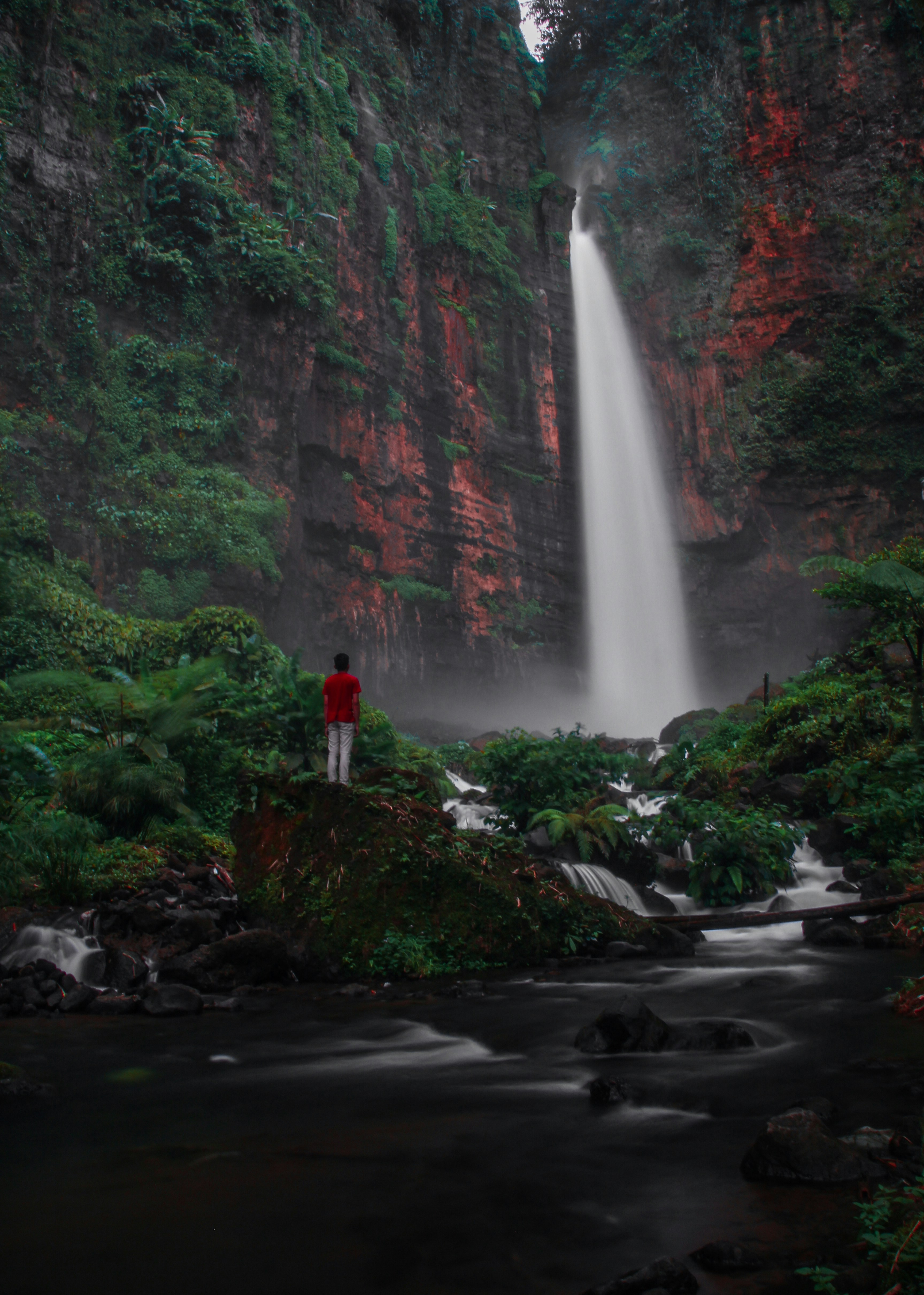 Person in a red jacket standing by a lush waterfall surrounded by verdant cliffs and flowing river.