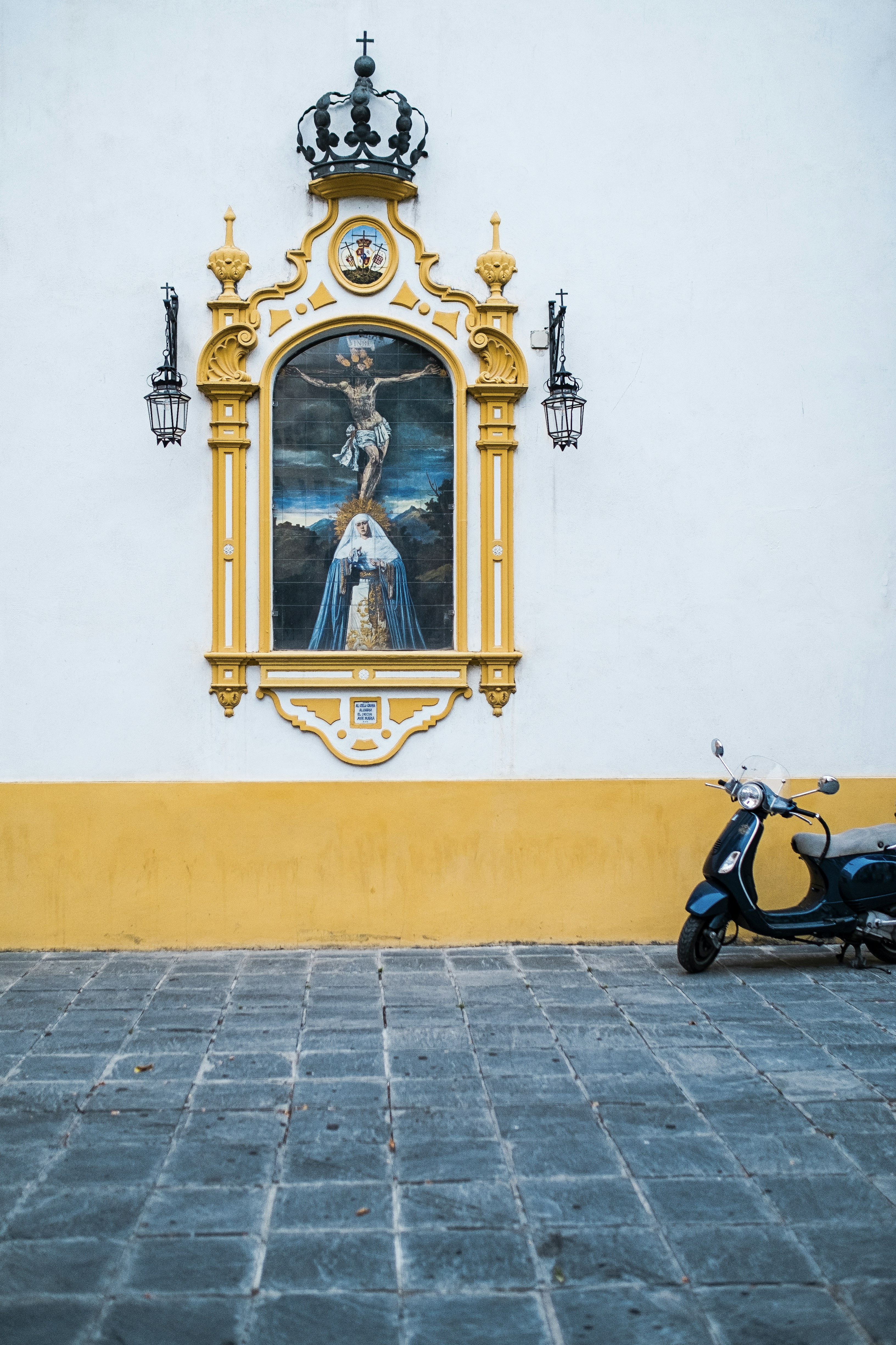 A striking mural of a religious figure framed in ornate yellow decor, complemented by a parked Vespa scooter on a cobblestone street.
