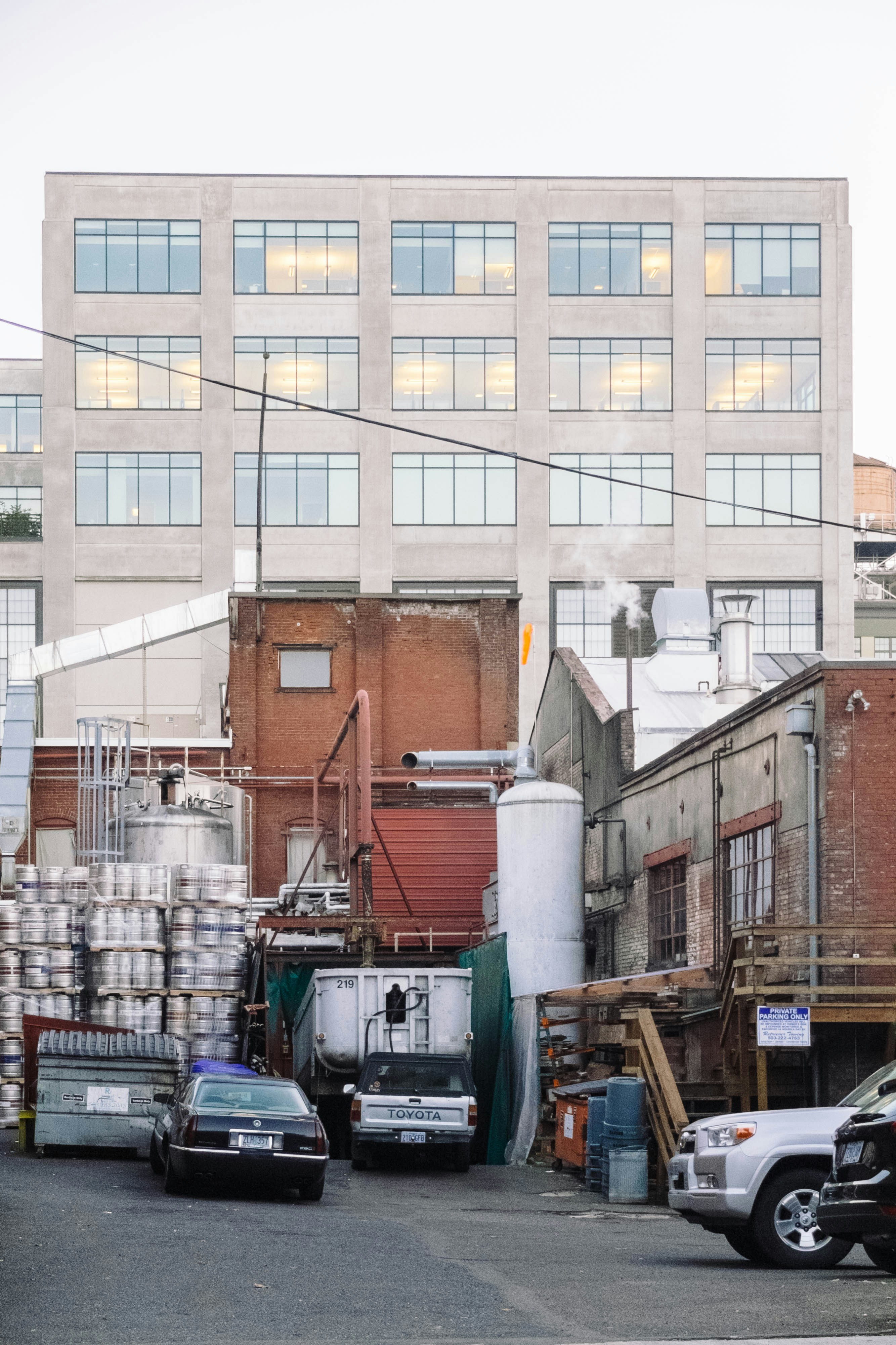 A view of a bustling urban alley showcasing vintage architecture and industrial elements, with parked vehicles and large storage tanks in the foreground.
