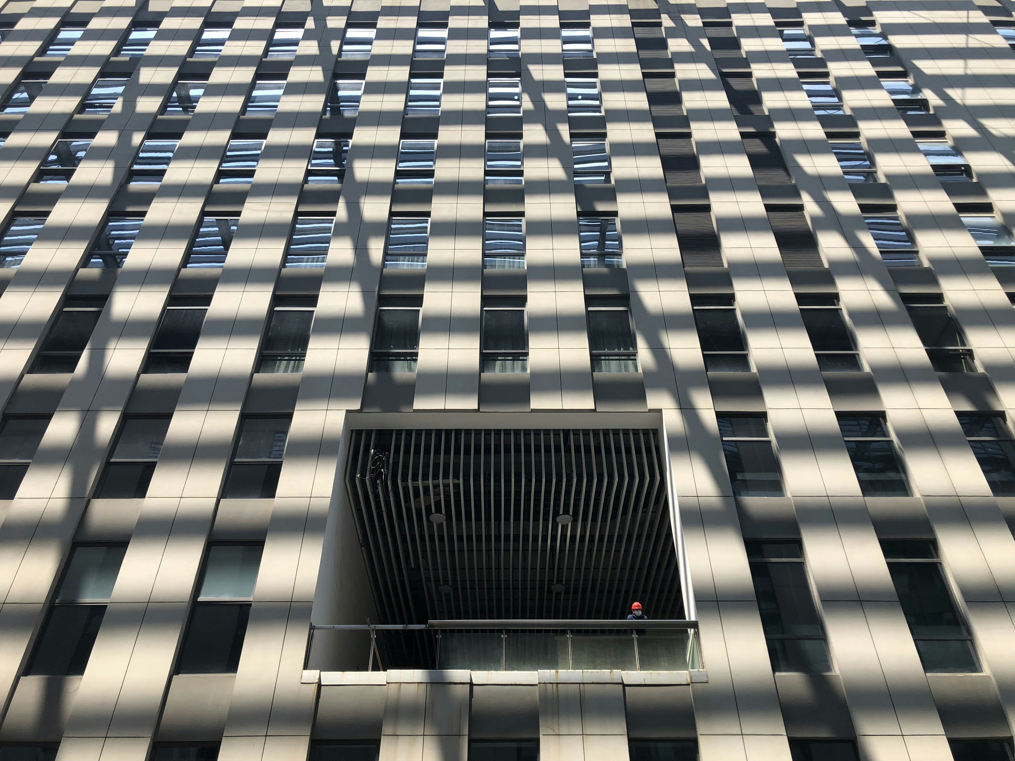 Sunlit glass facade with a checkered pattern of light and shadow, featuring a central balcony and a lone figure.