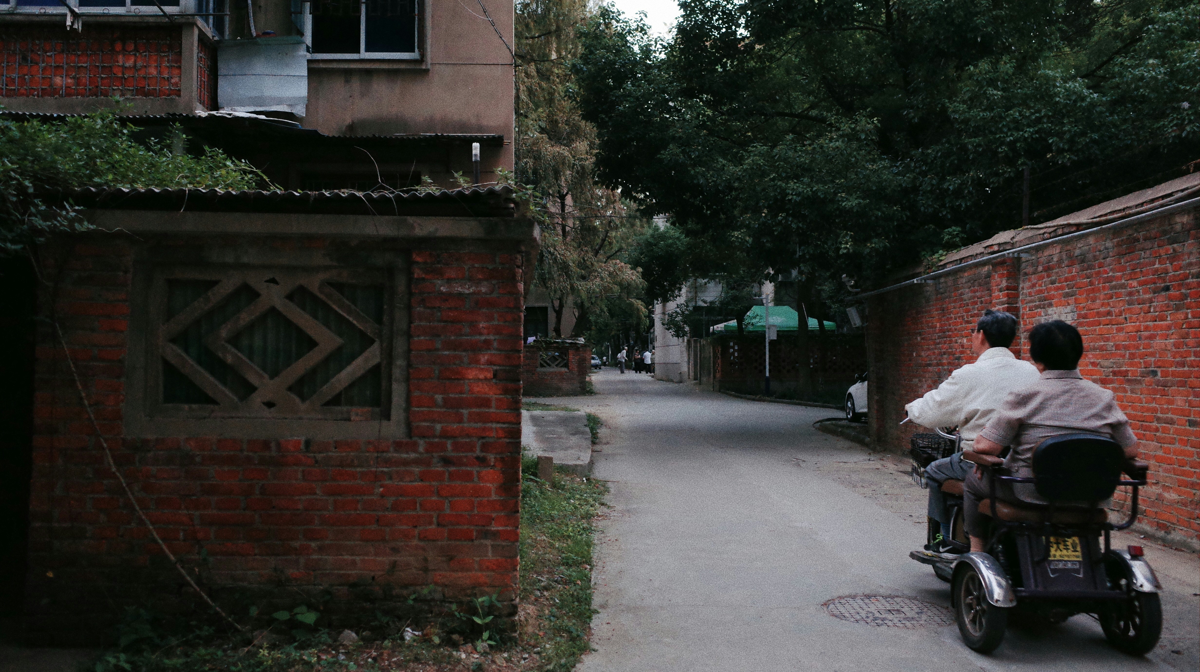 Two individuals on a scooter navigate a narrow alley lined with brick walls and greenery, evoking a sense of local life and exploration.