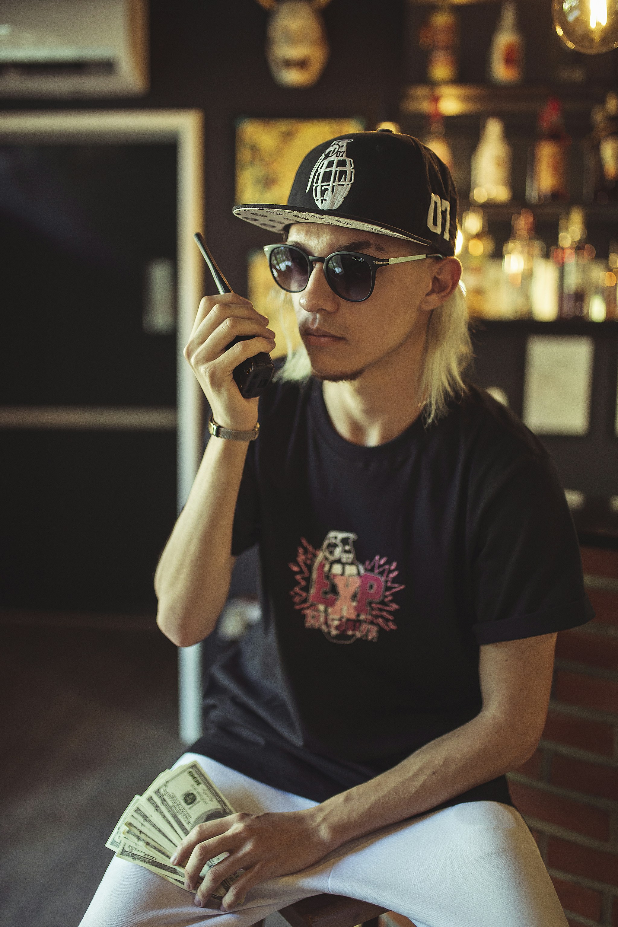 A young man in sunglasses and a cap holds a walkie-talkie while seated, displaying a stack of cash in his hand.