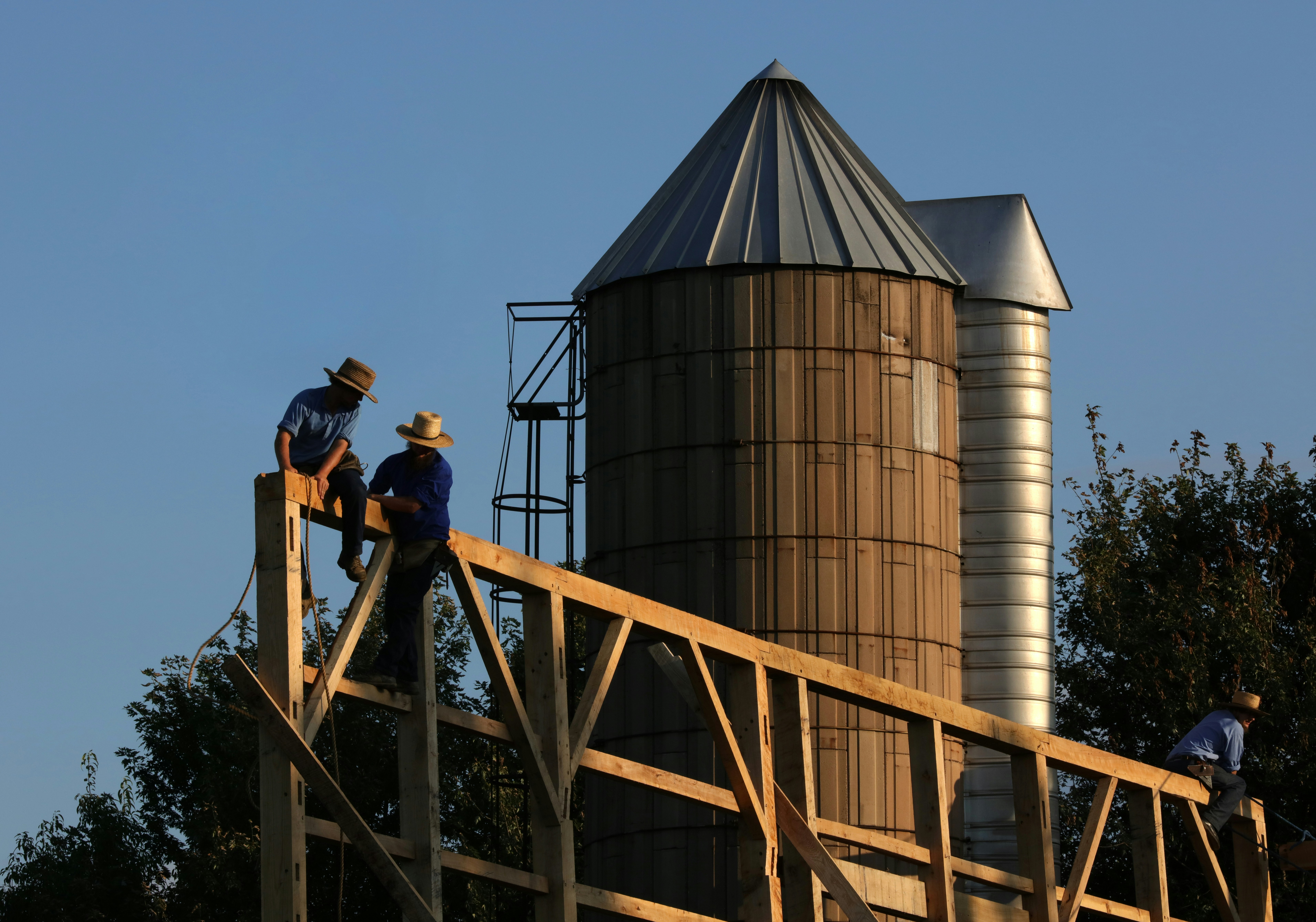 Two workers constructing a wooden frame under a clear sky, with a silos in the background. The scene captures the essence of agricultural labor and craftsmanship.