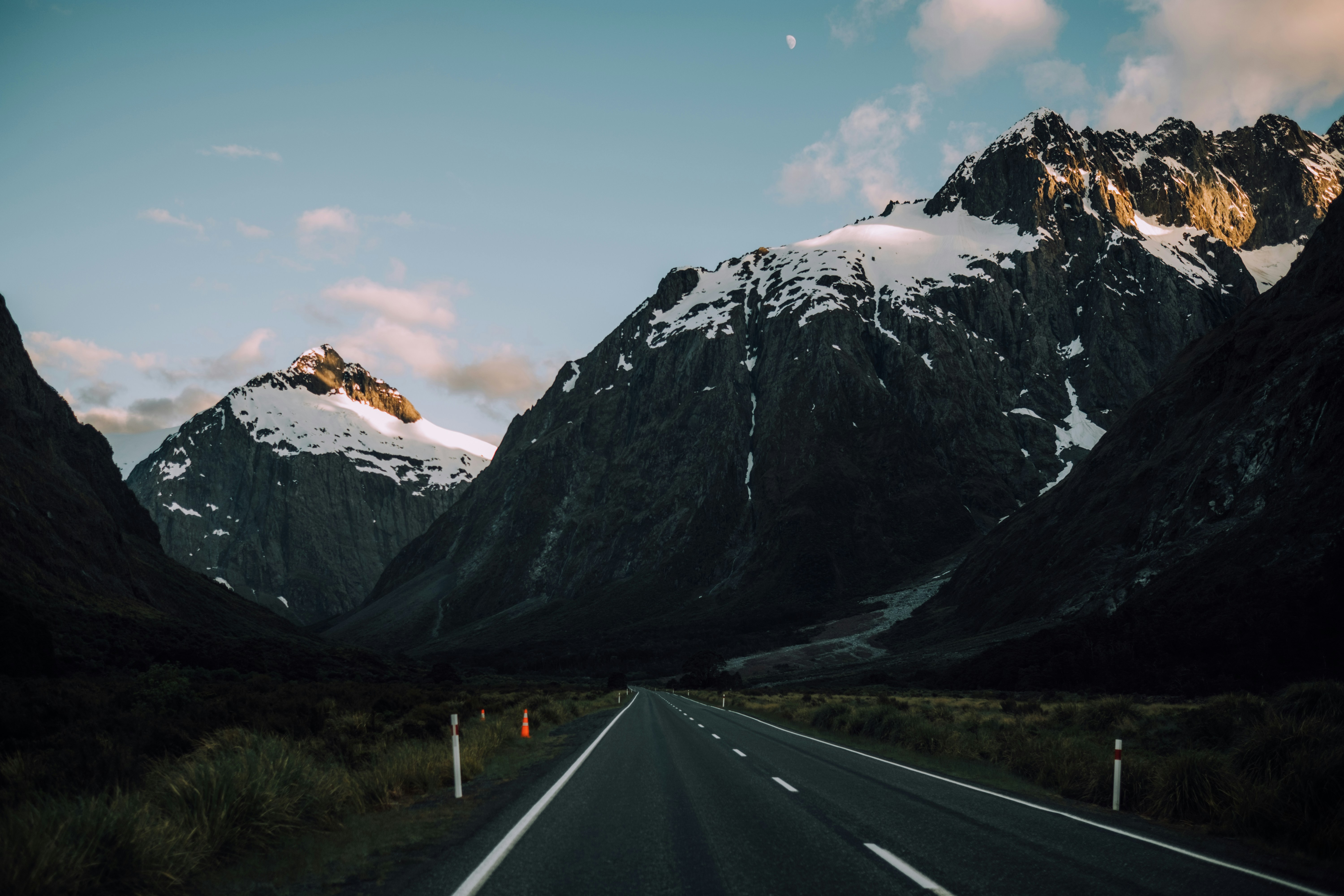 Wide road flanked by towering mountains with snow-capped peaks under a dusky sky.