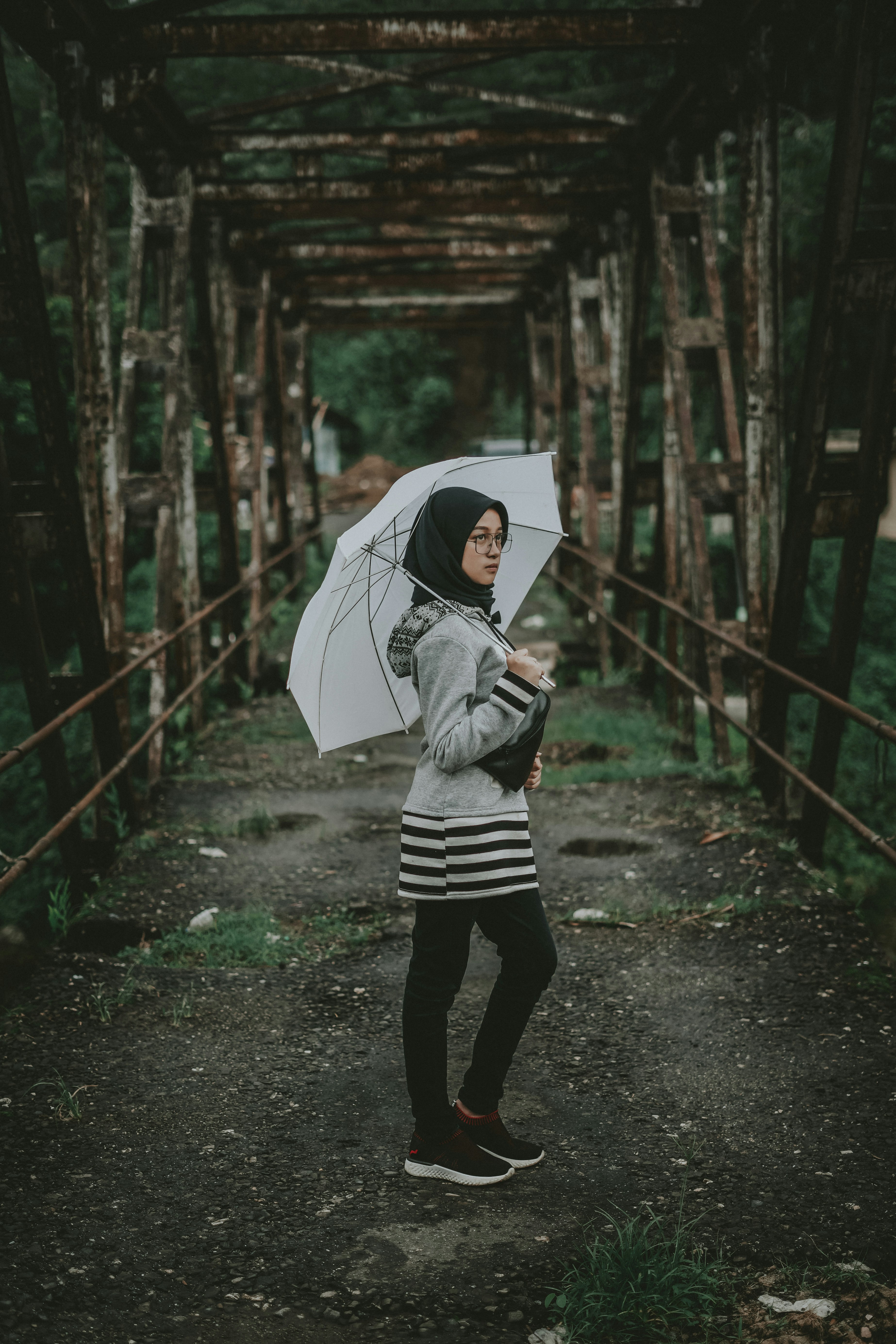 A woman stands gracefully on a rustic bridge, holding a white umbrella, surrounded by lush greenery and weathered metal structures.