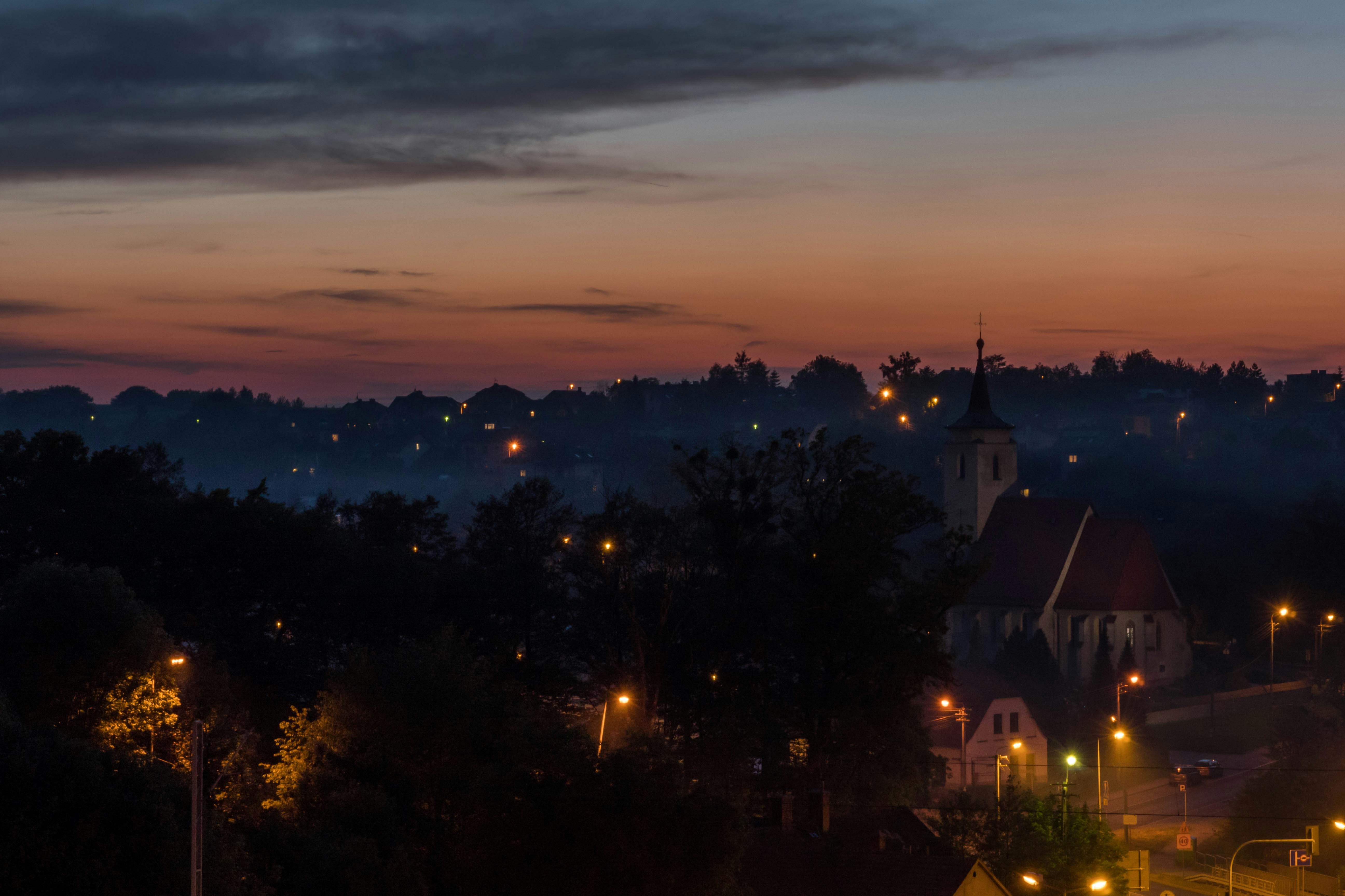 Dusk settles over a town with a church visible amidst glowing streetlights and silhouetted trees.