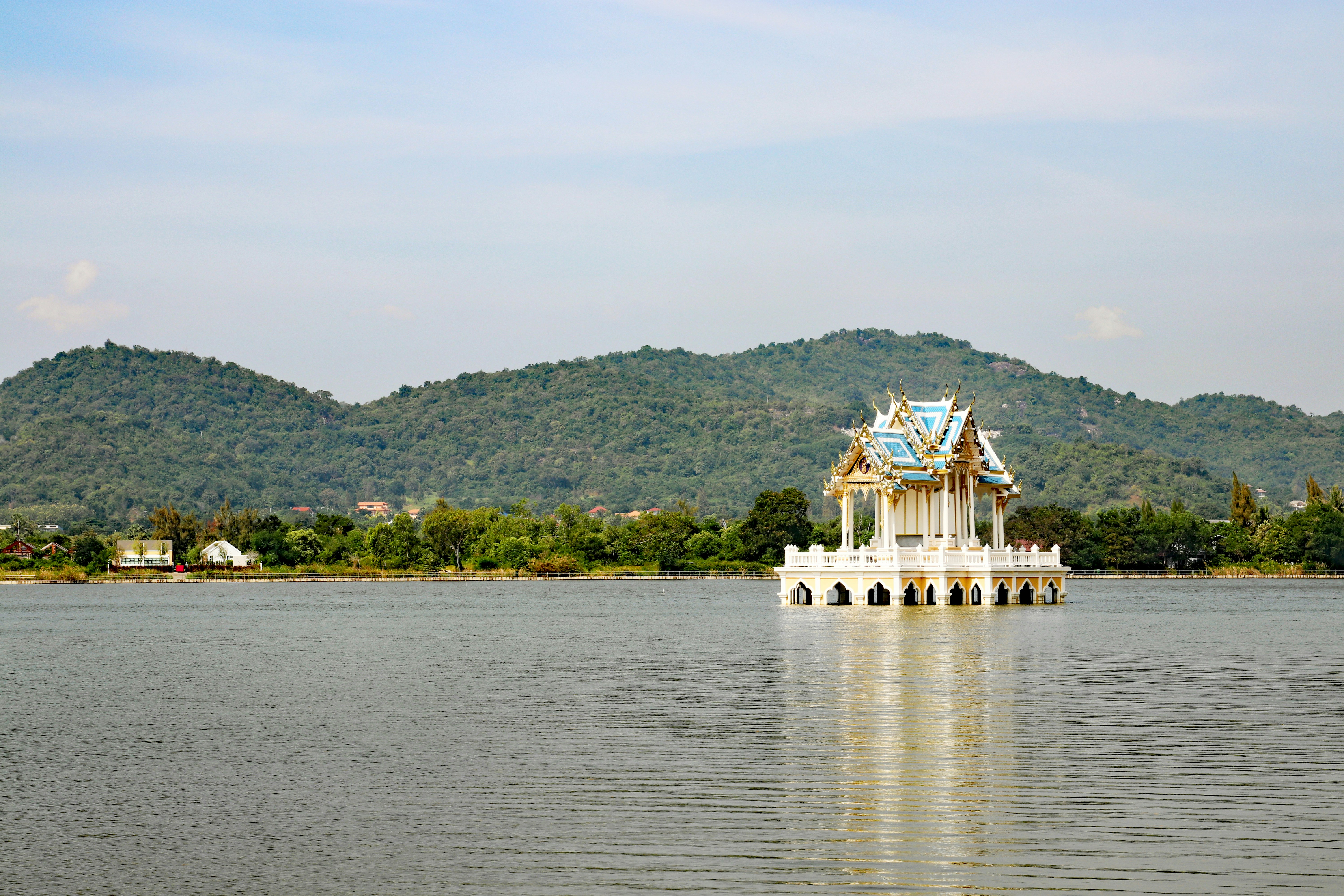 A beautifully crafted pavilion stands isolated on a tranquil lake, surrounded by lush green hills under a clear sky.