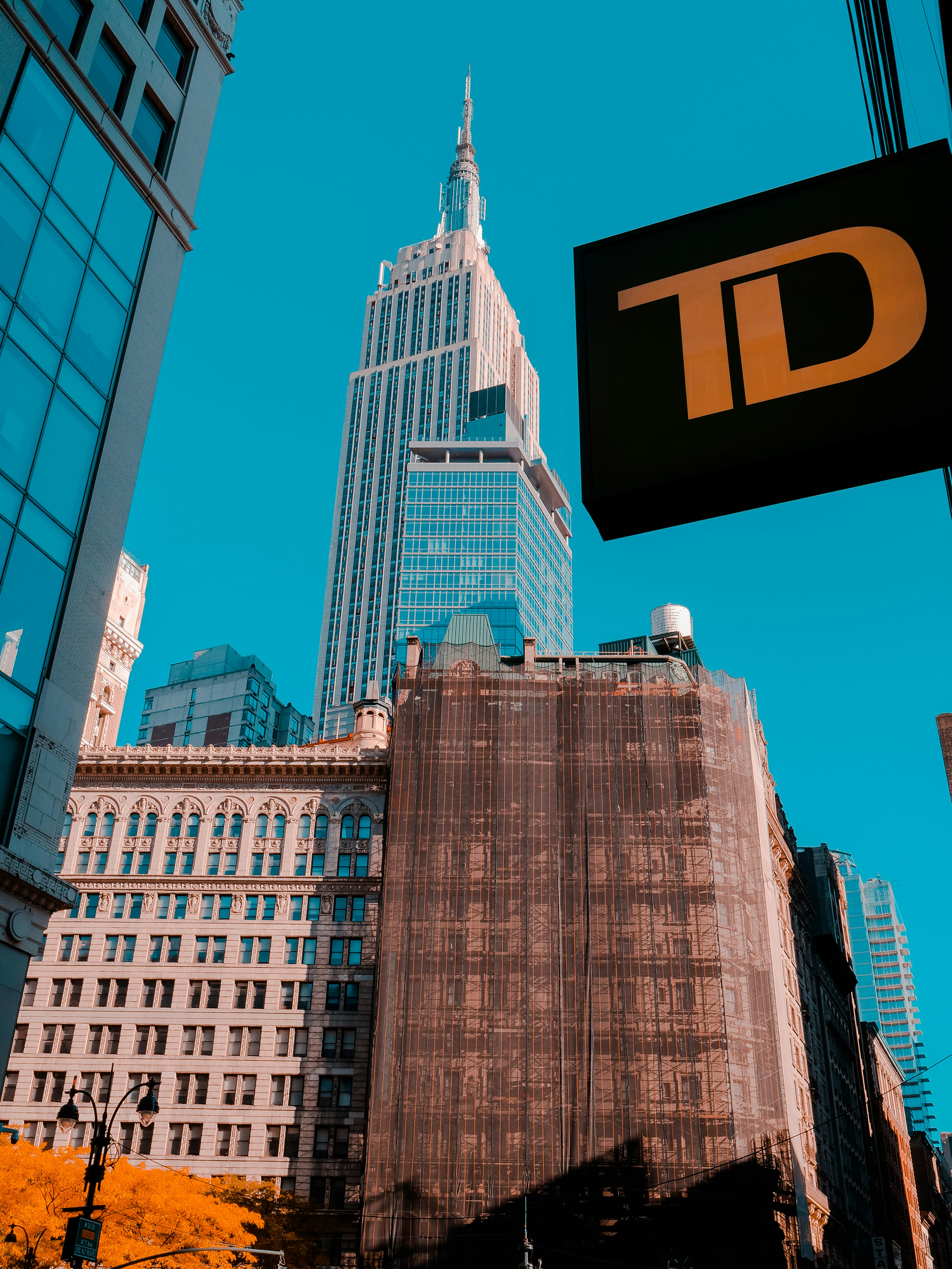 The Empire State Building towers above the cityscape, framed by modern architecture and a vibrant blue sky. A prominent TD sign adds a contemporary touch to the historic surroundings.