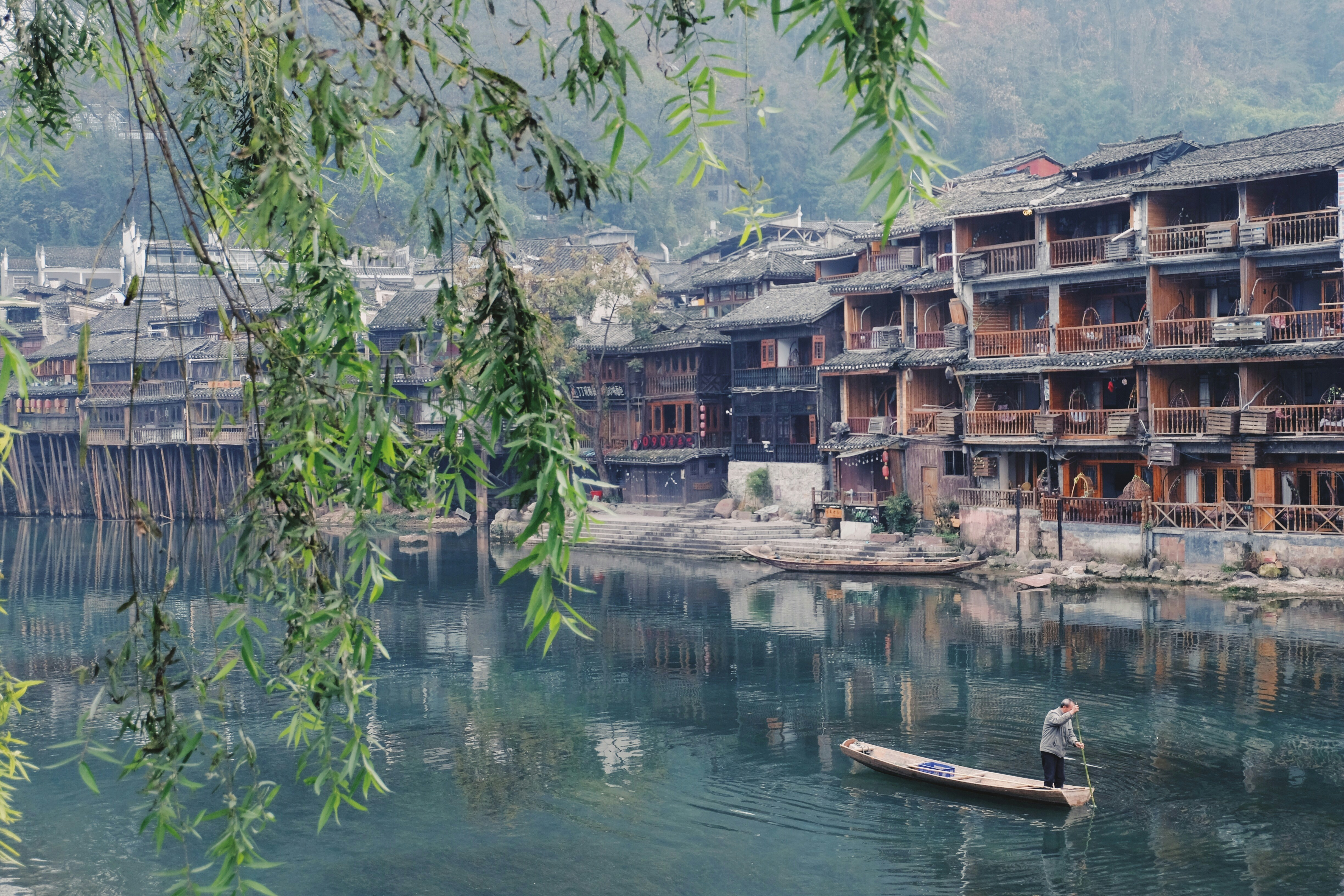 A lone fisherman navigates a tranquil river surrounded by traditional wooden buildings, framed by overhanging willow branches. The scene reflects a harmonious blend of nature and culture.