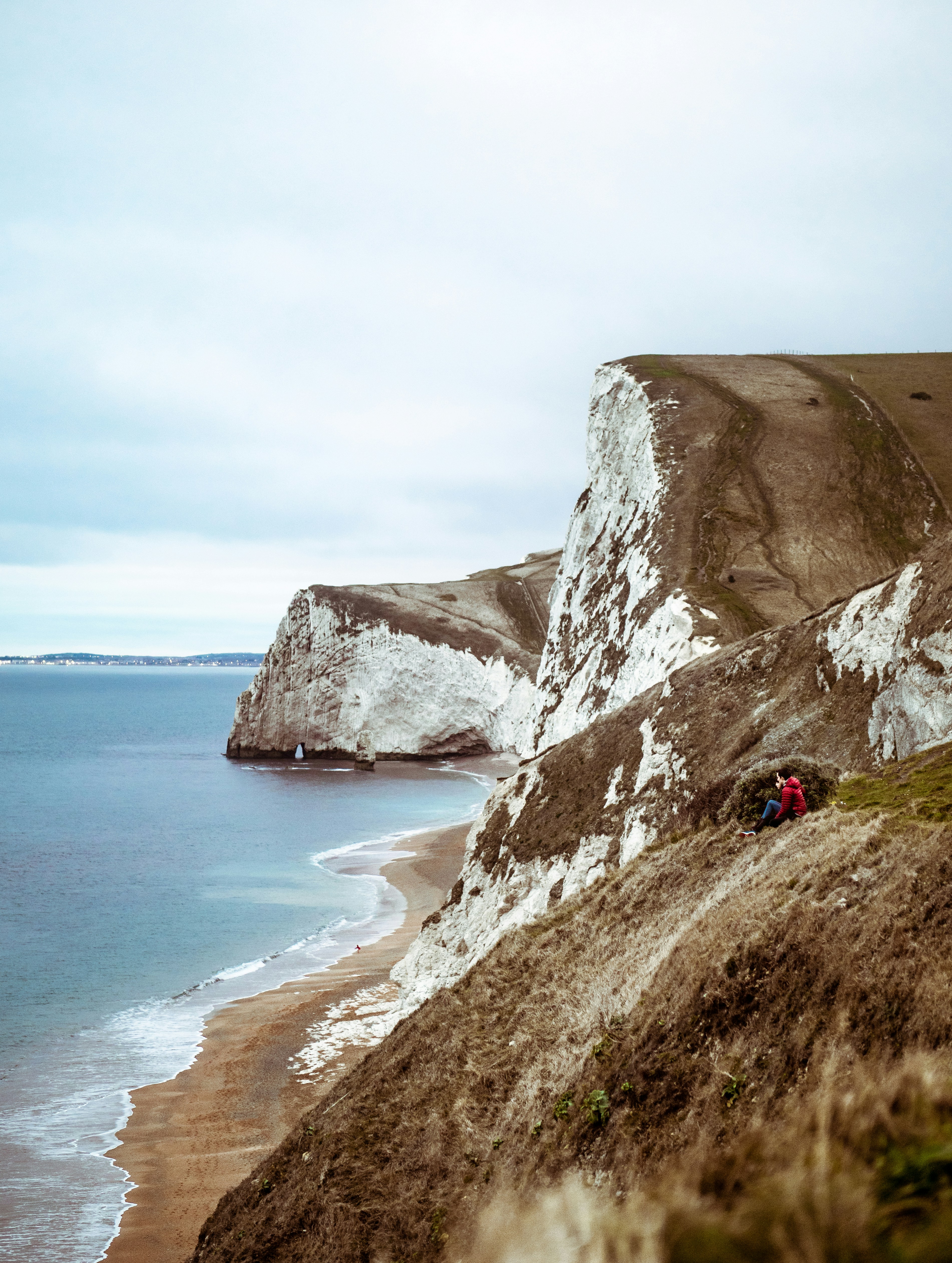 Two figures seated on a grassy cliff, overlooking a winding beach and rugged white cliffs under a cloudy sky.