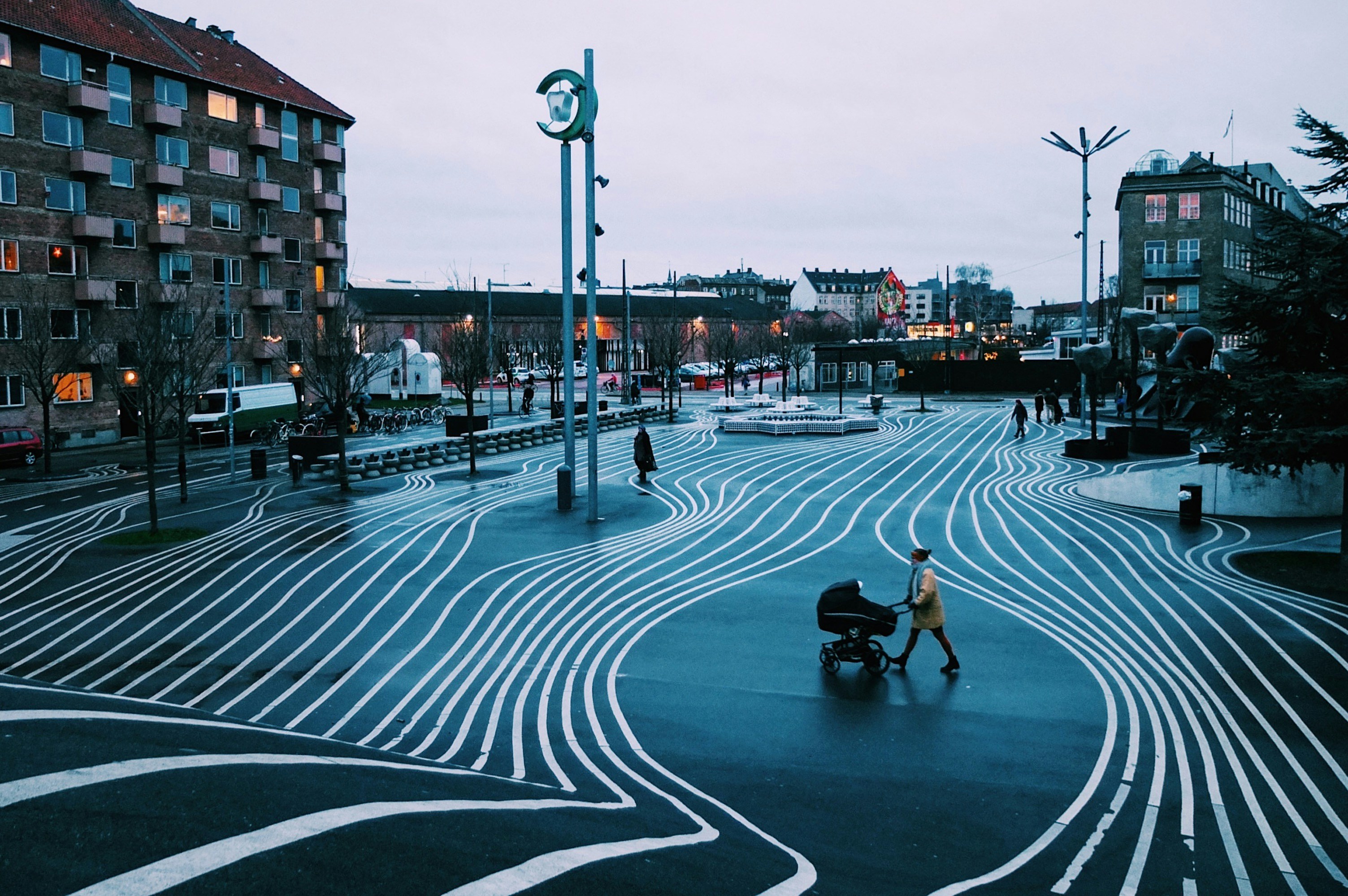 A pedestrian navigates a modern plaza marked by flowing white lines against a twilight backdrop, showcasing urban design and daily life.