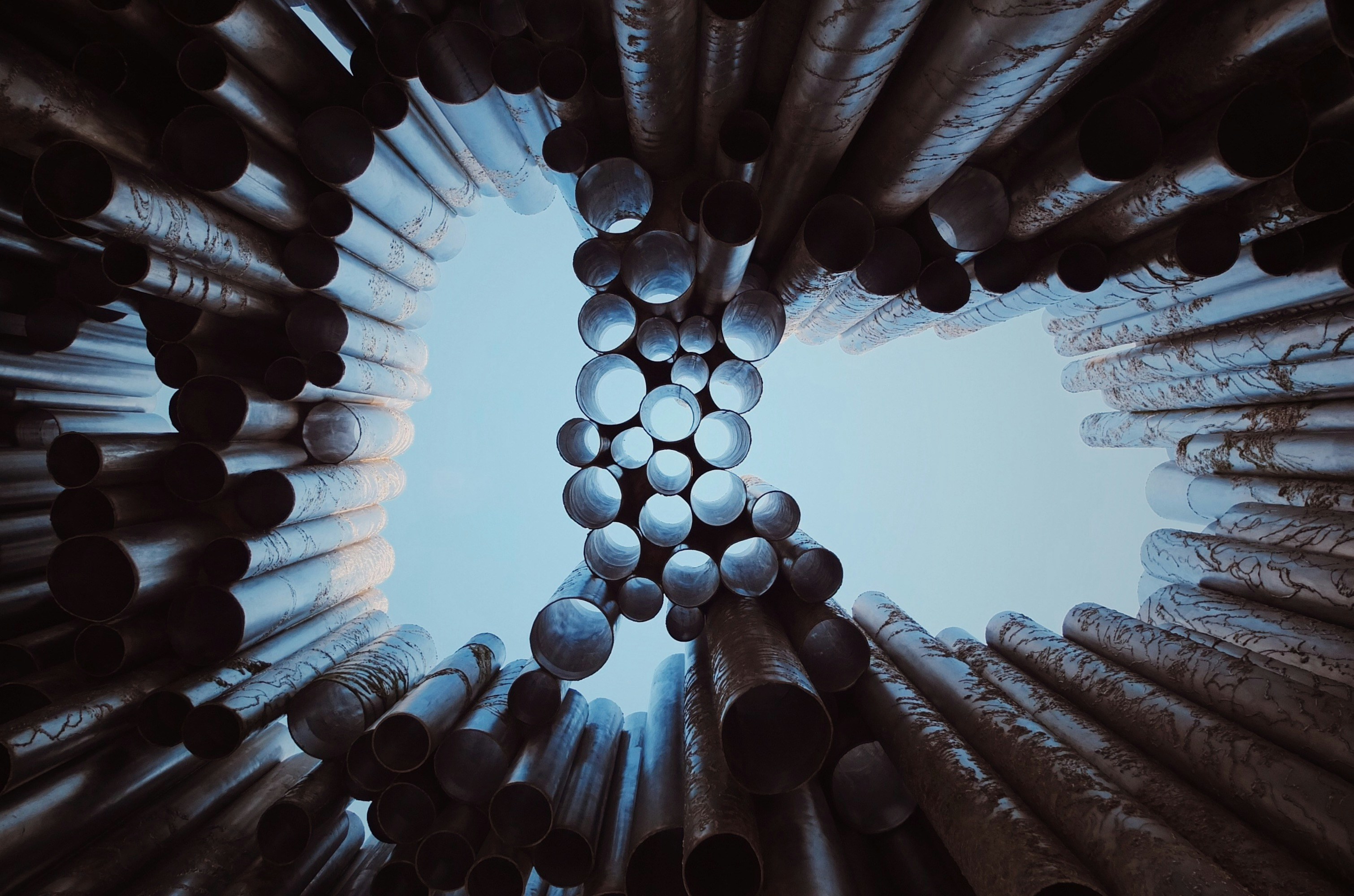 An abstract arrangement of cylindrical pipes converging towards a bright sky, creating a unique visual perspective. The interplay of shapes and light draws the eye upward.